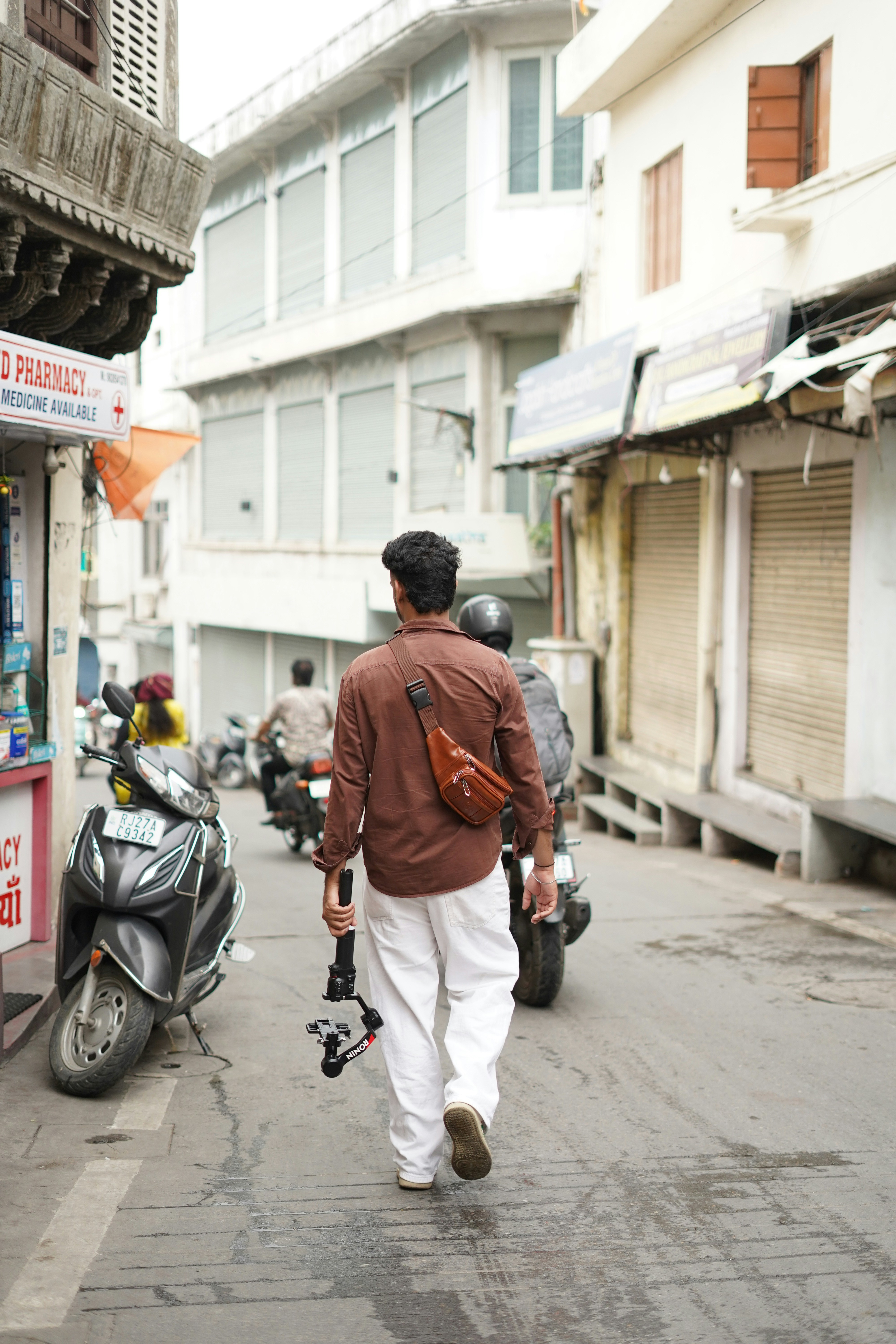 A man walks down a narrow street, camera in hand, flanked by parked scooters and shuttered shops. The scene captures the essence of urban exploration.