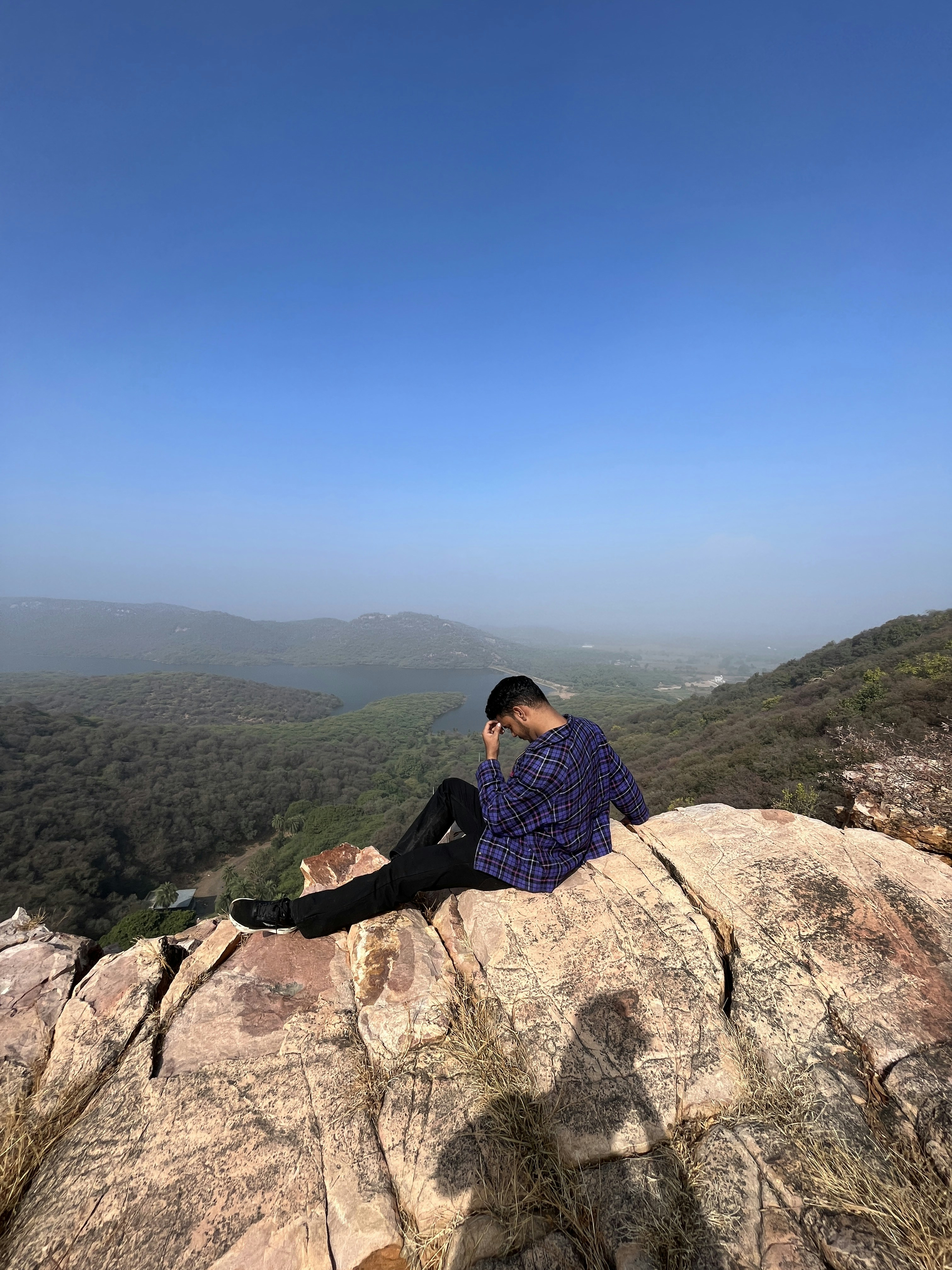 A person seated on a rocky outcrop, gazing thoughtfully into the vast green valley below. The clear blue sky enhances the serene atmosphere.
