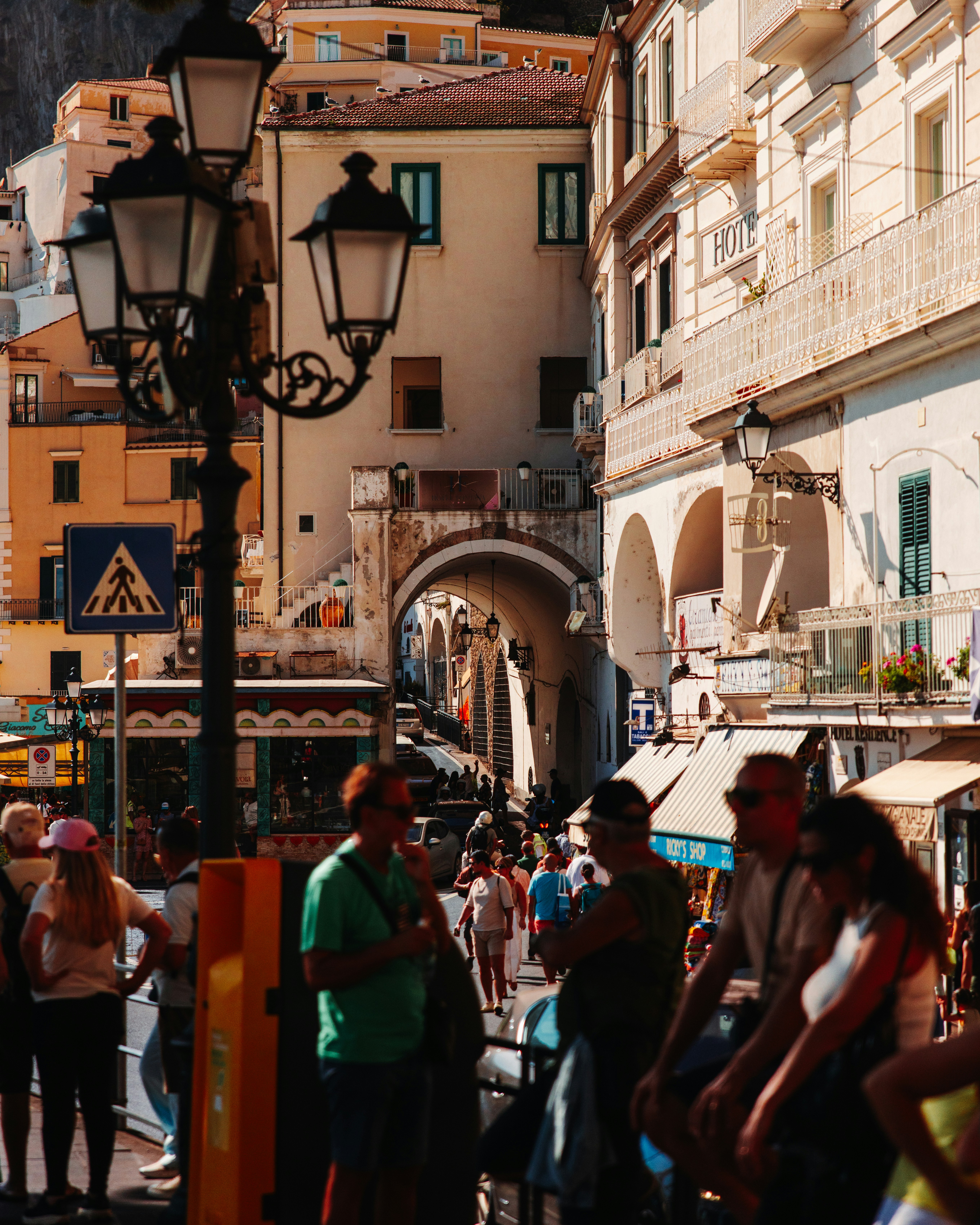 Bustling street scene in a coastal town, featuring a mix of architecture and lively pedestrians. A vintage lamp post adds character to the foreground.