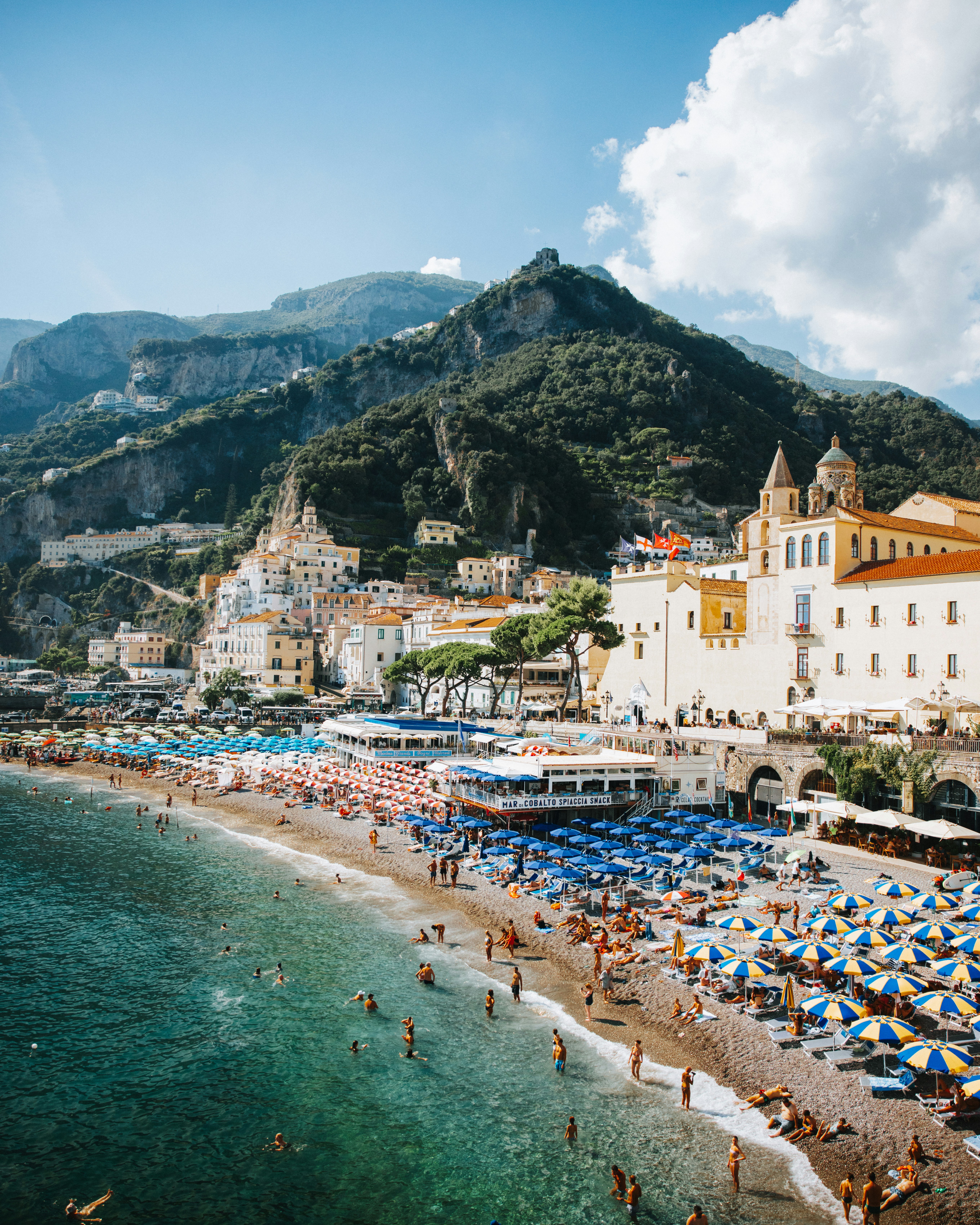 Crowded beach with umbrellas and buildings on a sunny day.