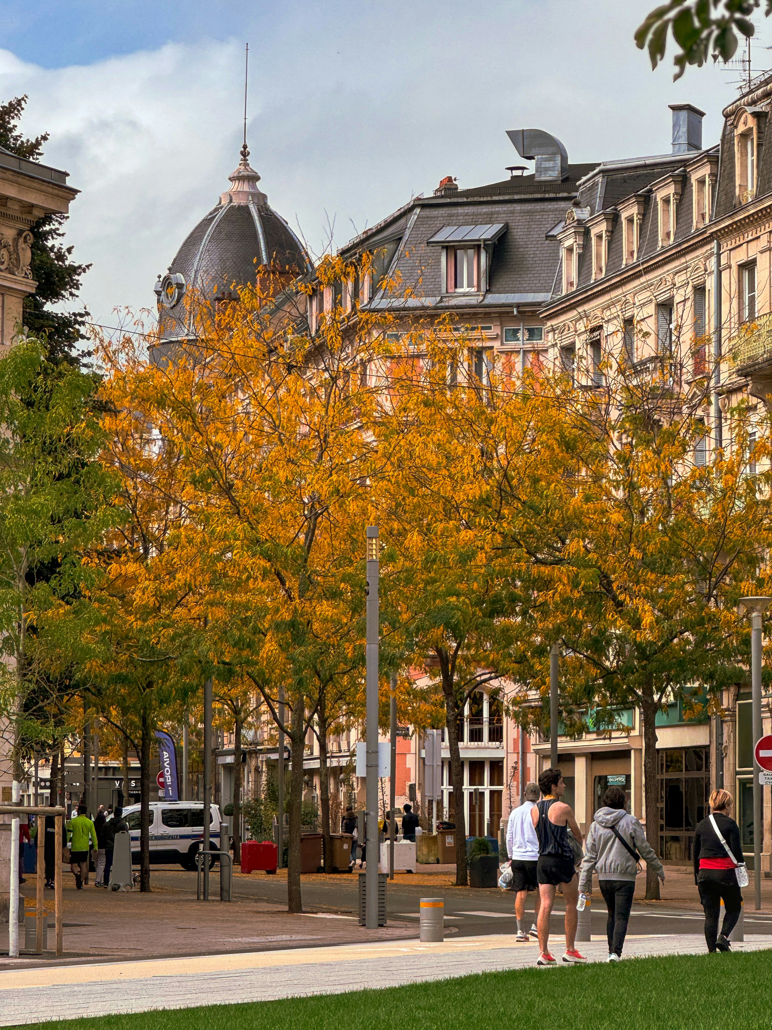 Teintes automnales 🍁 | People walk past trees with autumn foliage in a city.