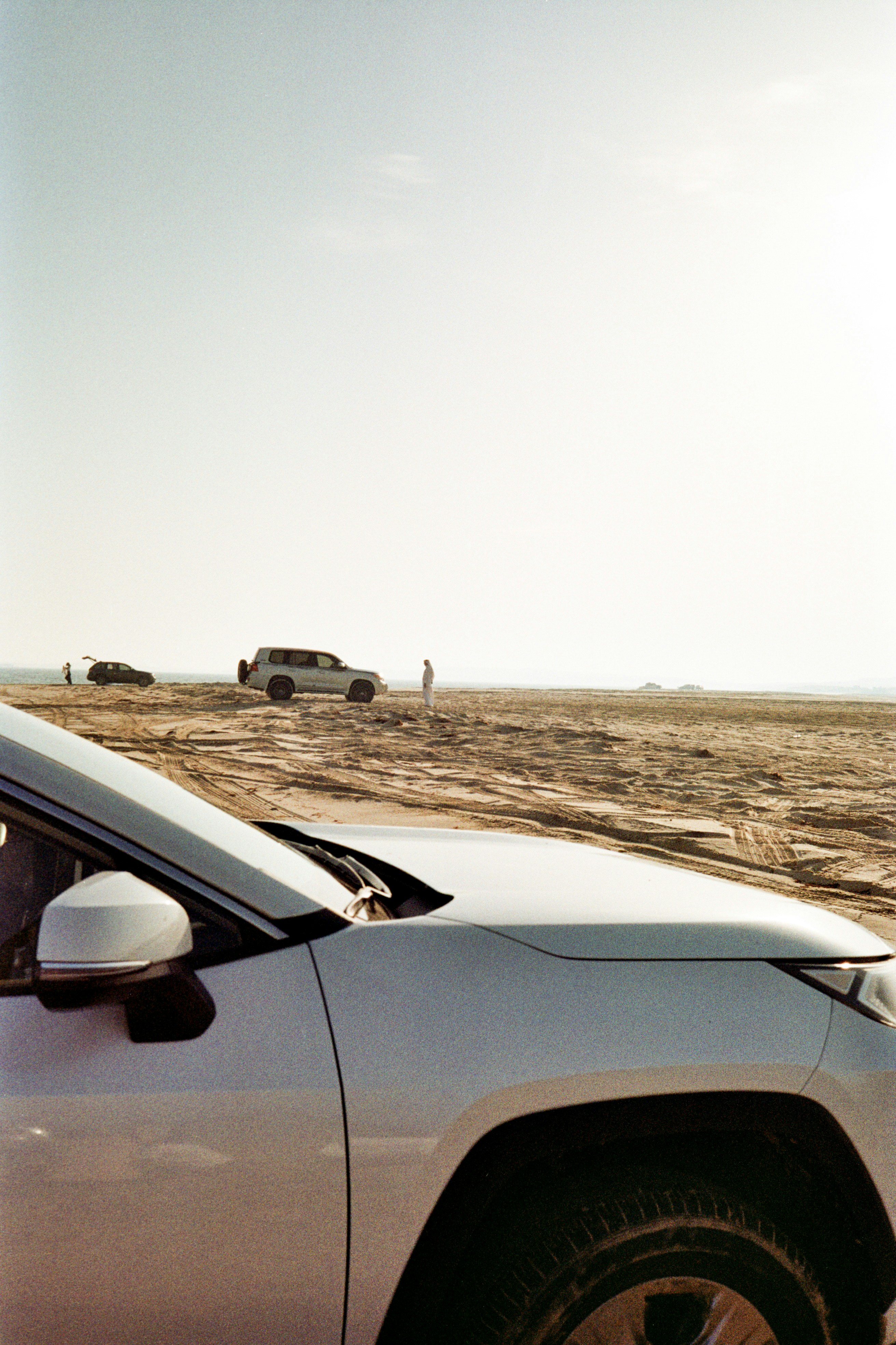 A silver SUV foregrounded against a vast desert landscape, with distant vehicles and a lone figure exploring the terrain.