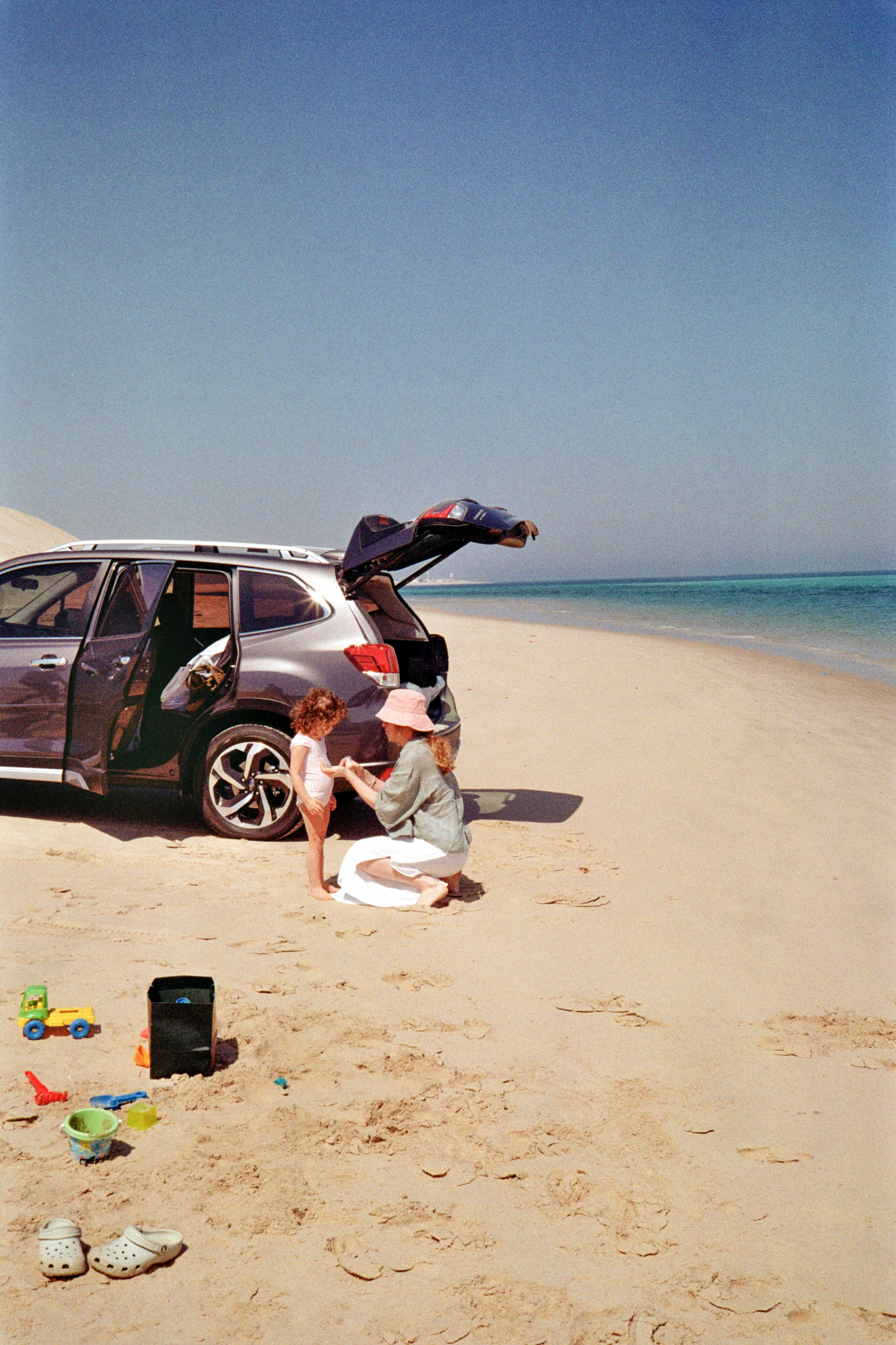 A child plays on the beach while an adult assists nearby, with a car parked in the background. Colorful beach toys are scattered in the sand.