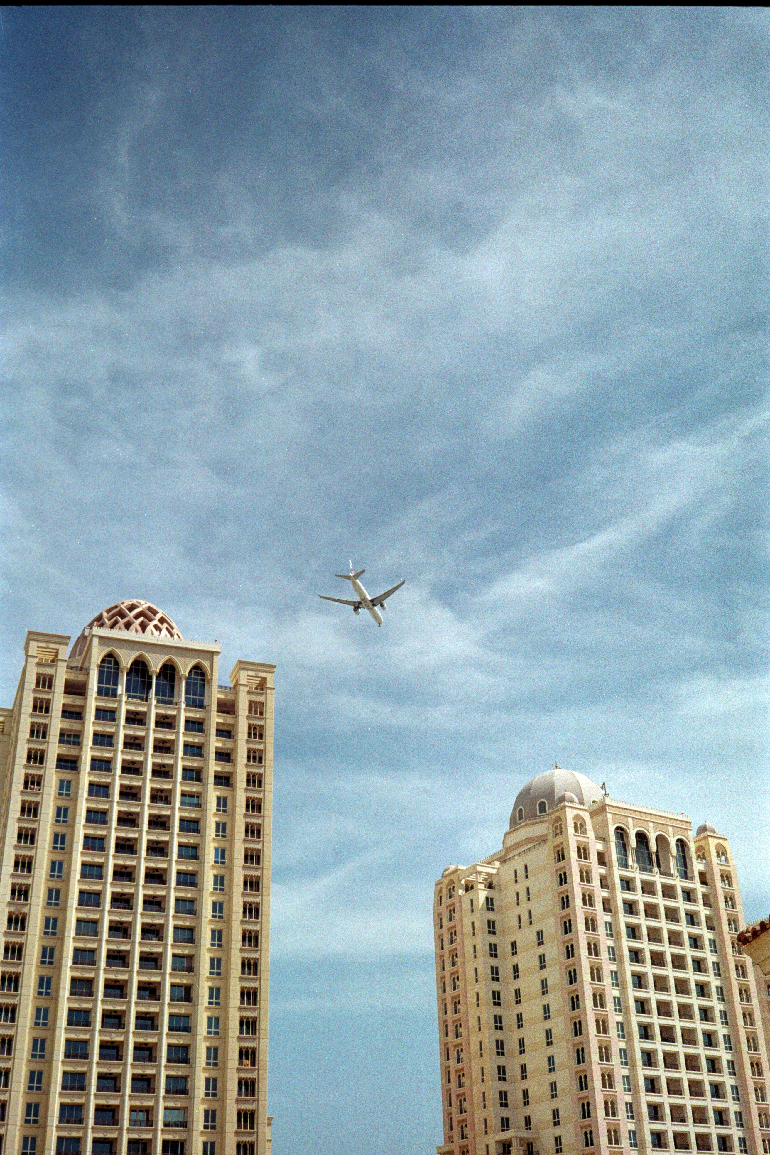 Airplane soaring above modern skyscrapers under a bright blue sky with wispy clouds.