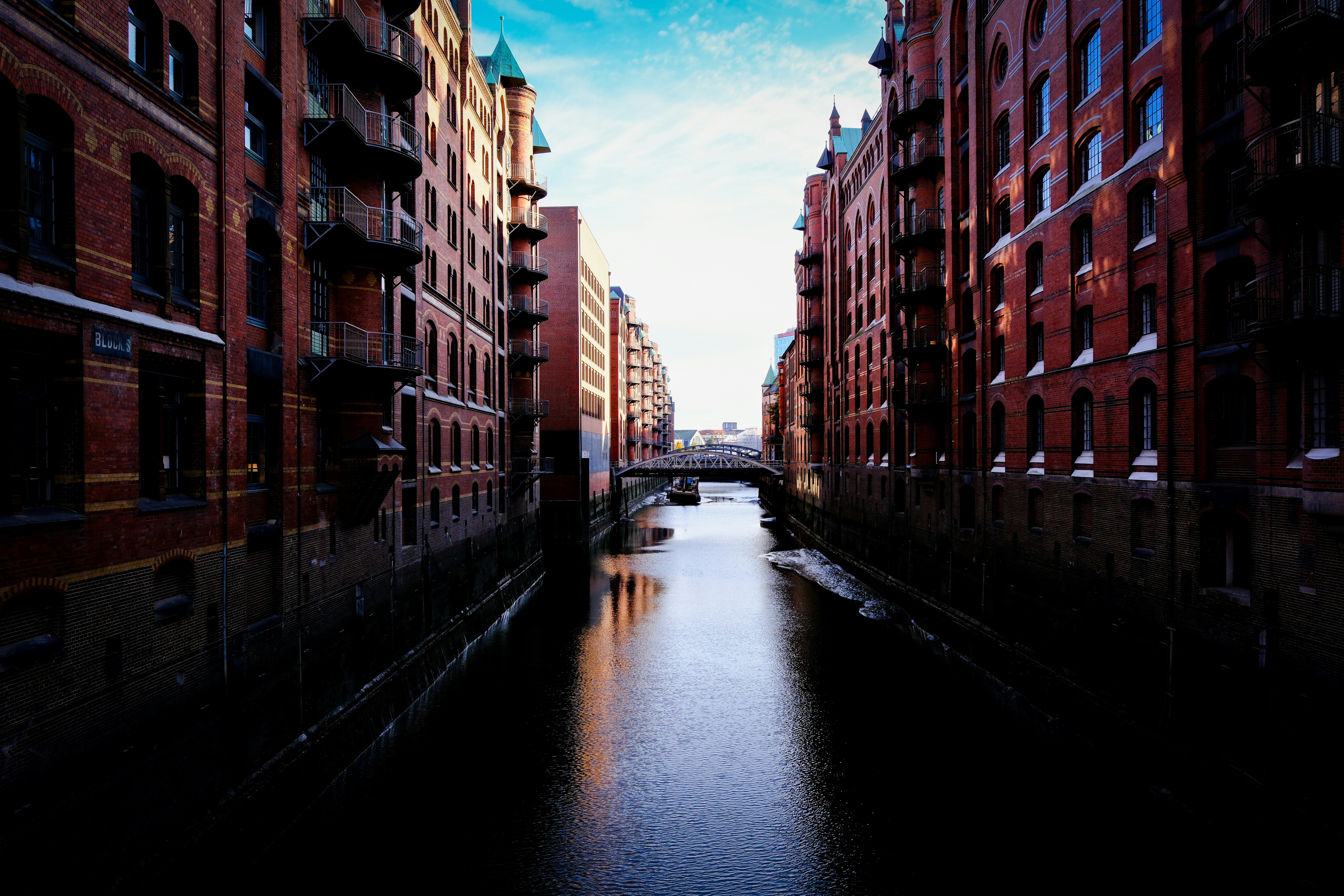 Historic red-brick warehouses lining a tranquil canal, reflecting the sky and architecture in the water. A bridge connects the two sides, inviting exploration.