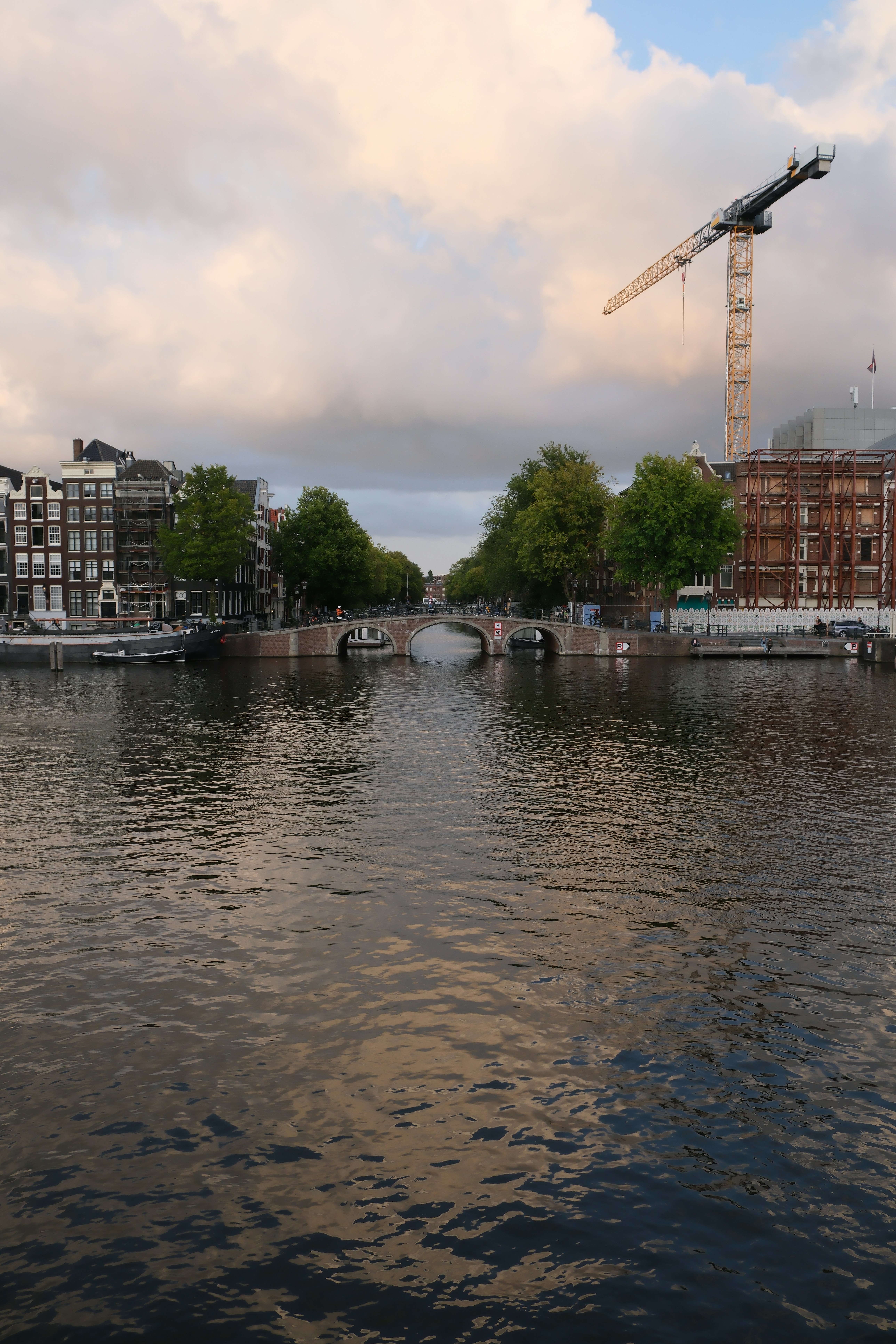Historic canal scene in Amsterdam featuring a charming bridge, lush trees, and a construction crane, symbolizing the city's blend of tradition and modernity.
