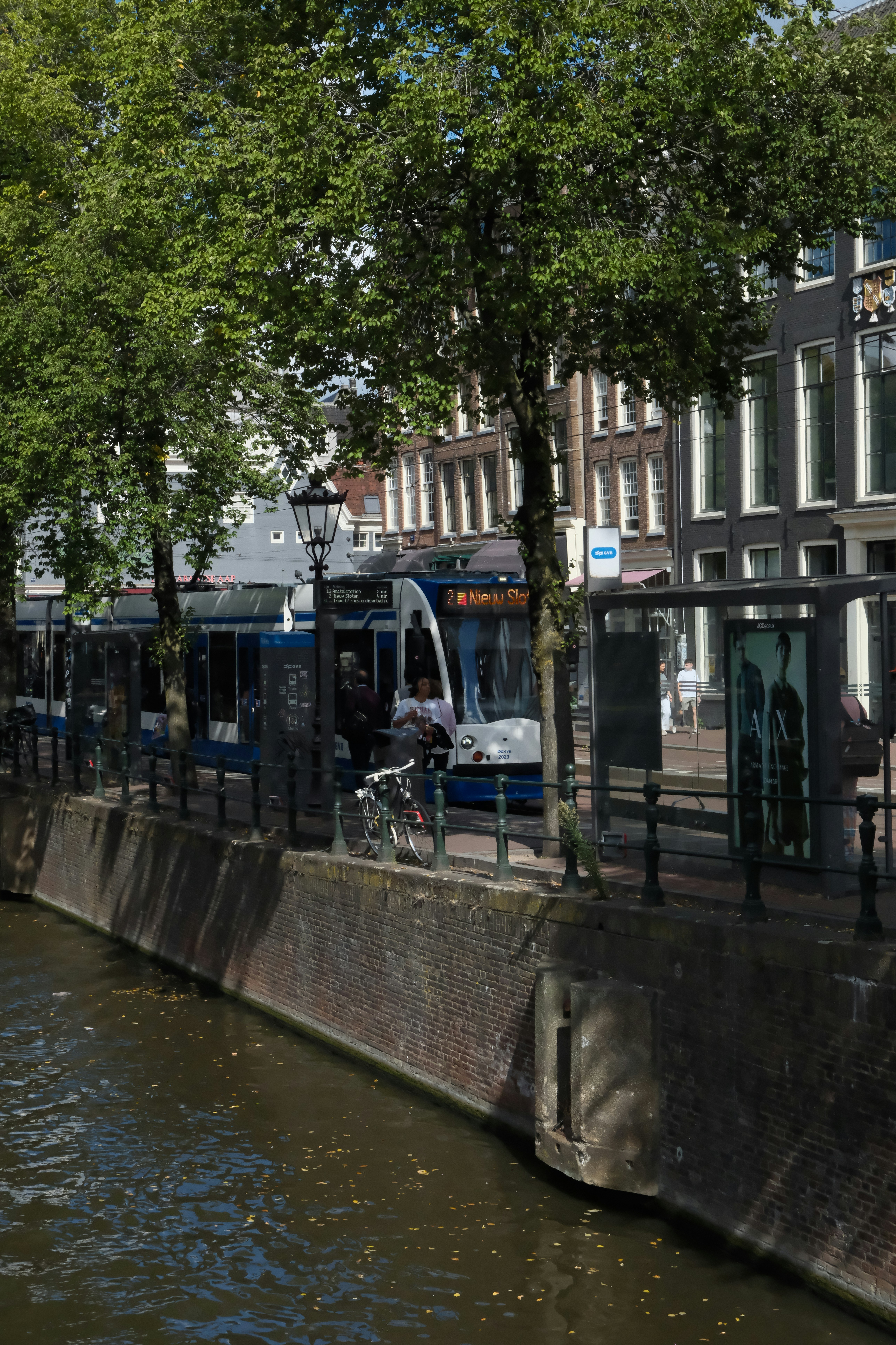 Tram navigating alongside a canal, framed by lush trees and historic architecture. A bicycle leans against the railing, adding to the urban charm.