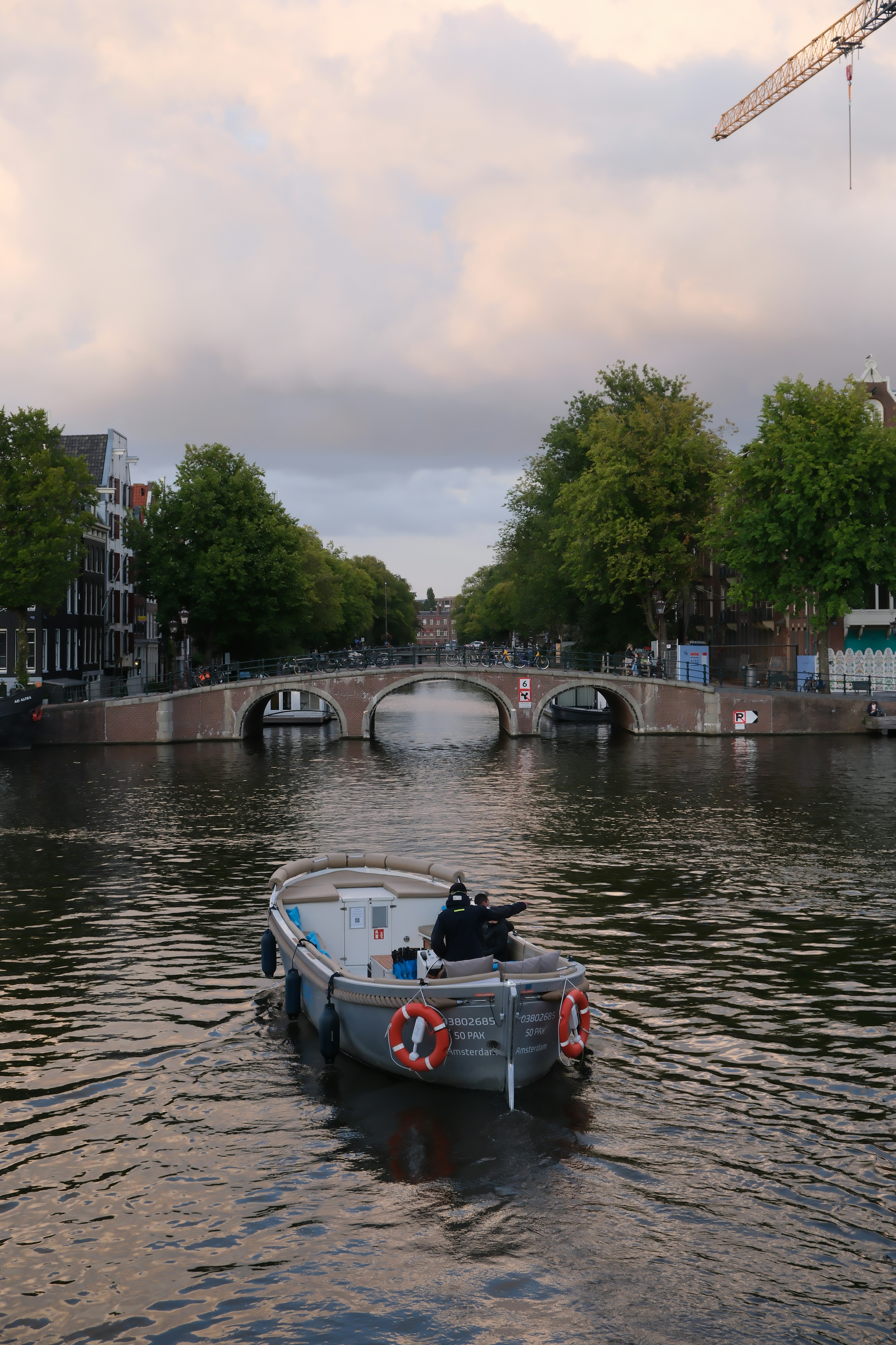 A small boat navigates the calm waters of an Amsterdam canal, framed by lush trees and historic architecture under a softly lit sky.