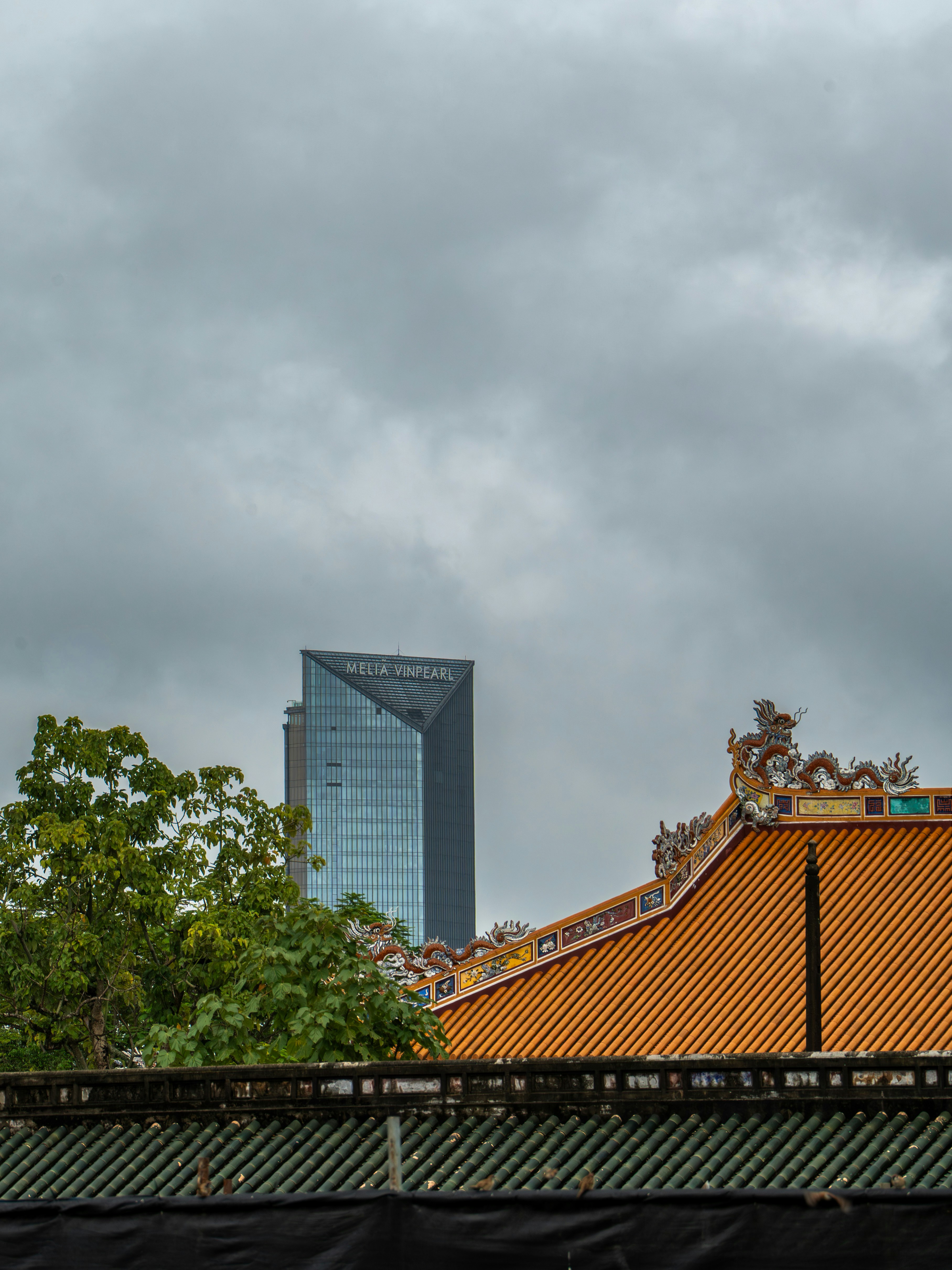 A modern skyscraper rises behind the traditional tiled rooftops of the Imperial City in Huế, Vietnam. The contrast between contemporary architecture and ancient design highlights the blend of history and modern development in Vietnam’s former imperial capital.