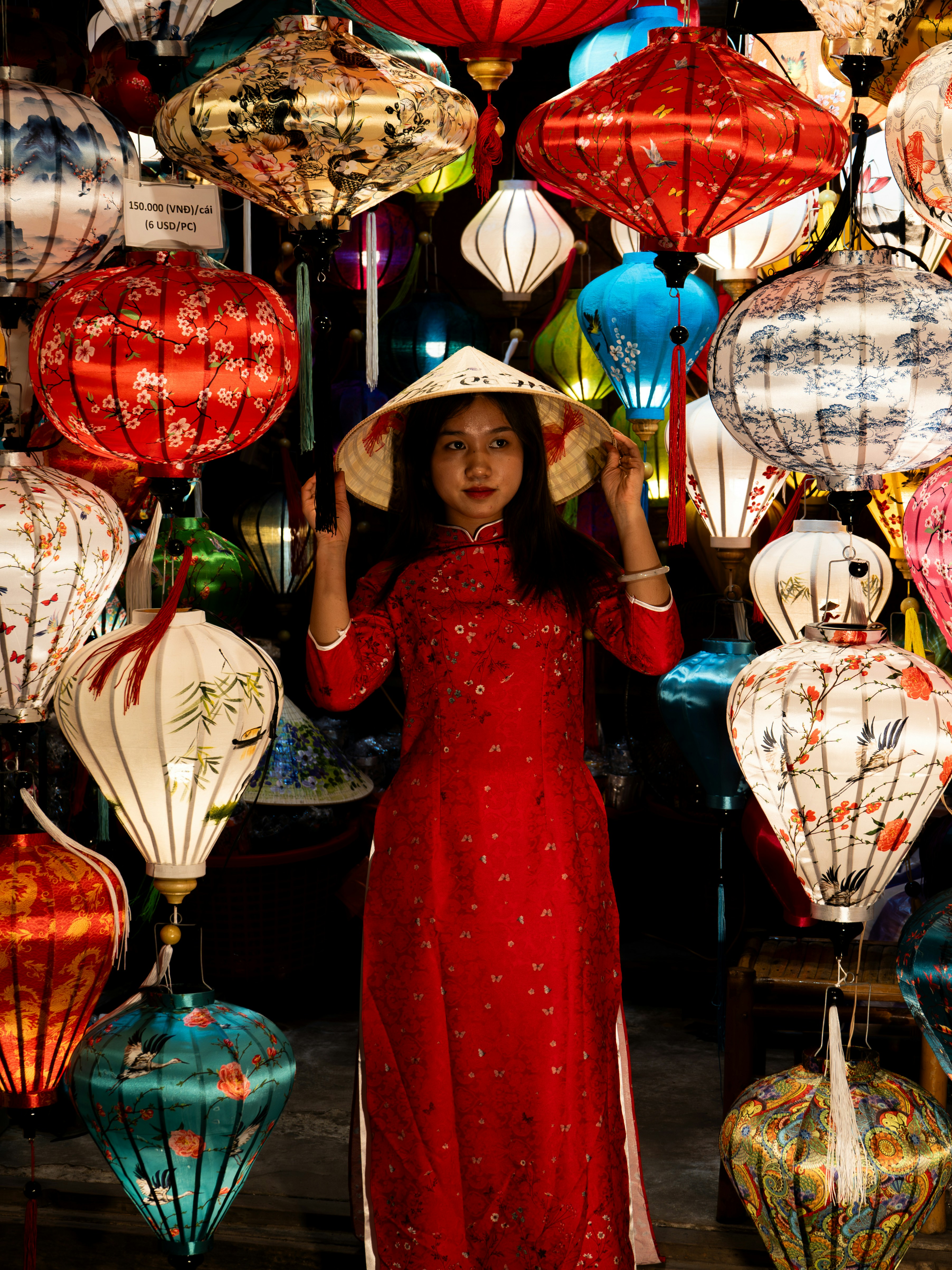 A woman wearing a traditional red áo dài and conical hat stands among colorful lanterns in Hoi An, Vietnam. The vibrant lights and traditional attire highlight the cultural beauty and timeless charm of Hoi An’s famous lantern market.