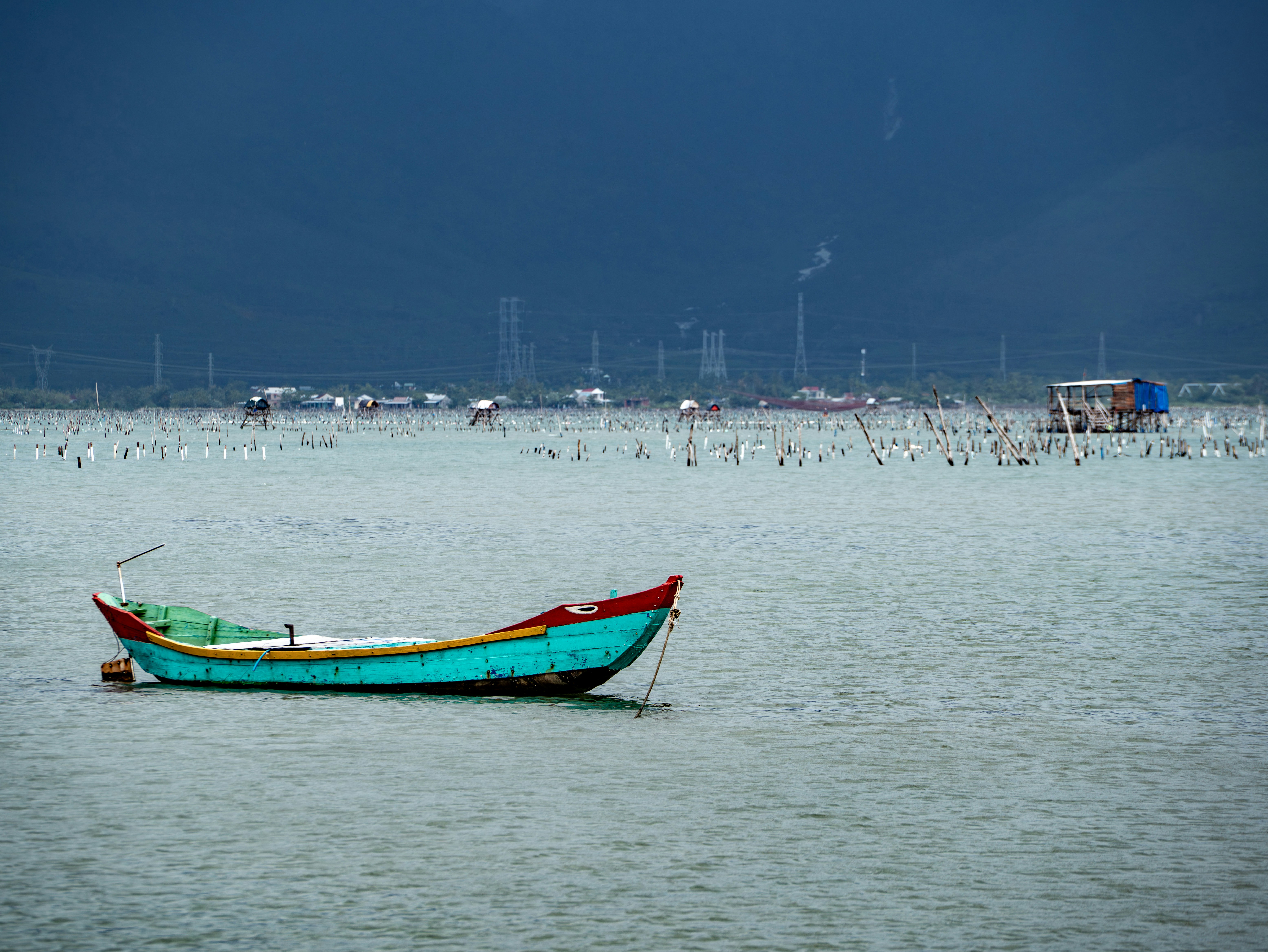 Colorful fishing boat anchored in a tranquil body of water, surrounded by distant structures and a moody sky.