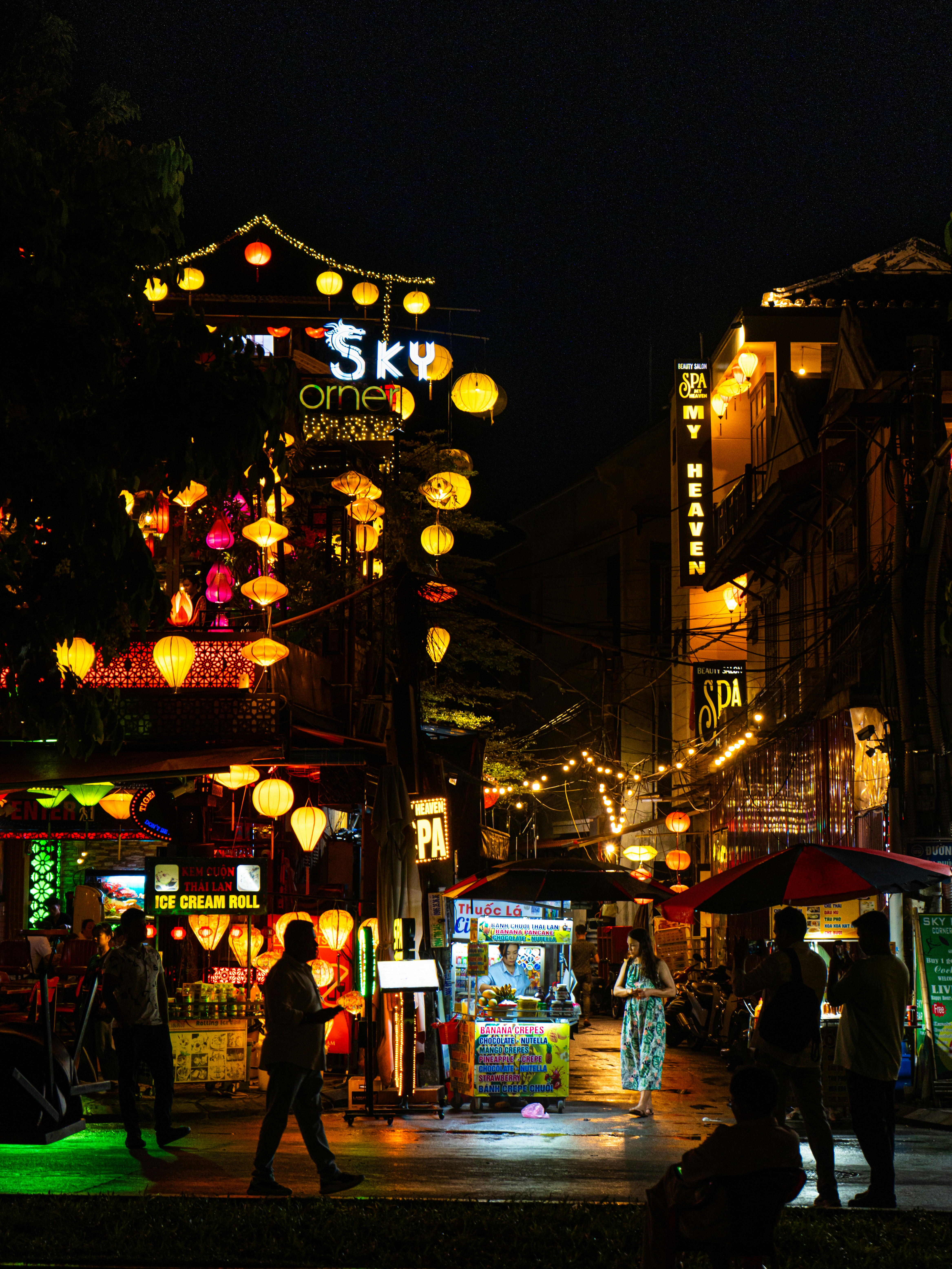 A vibrant night scene in Hoi An, Vietnam, filled with colorful lanterns, neon lights, and street vendors. The lively streets capture the city’s energetic nightlife and the blend of tradition and modern charm that defines Hoi An after dark. | Street scene at night with glowing lanterns and shops.