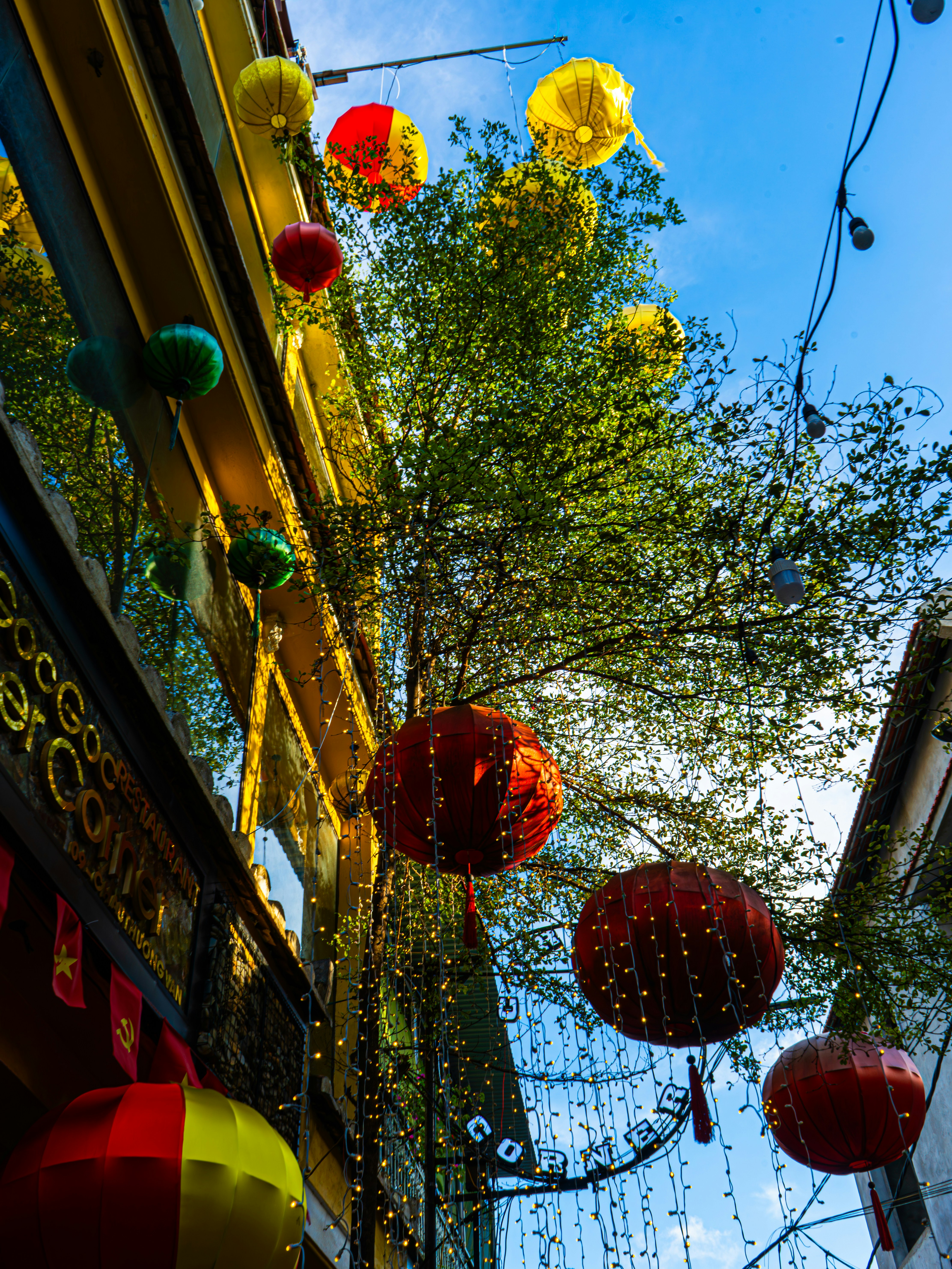 Colorful lanterns hang above a narrow street in Hoi An, Vietnam, glowing under the evening sunlight. The red and yellow lanterns, paired with decorative string lights, capture the festive and vibrant spirit of Hoi An’s ancient town and its traditional Vietnamese architecture.
