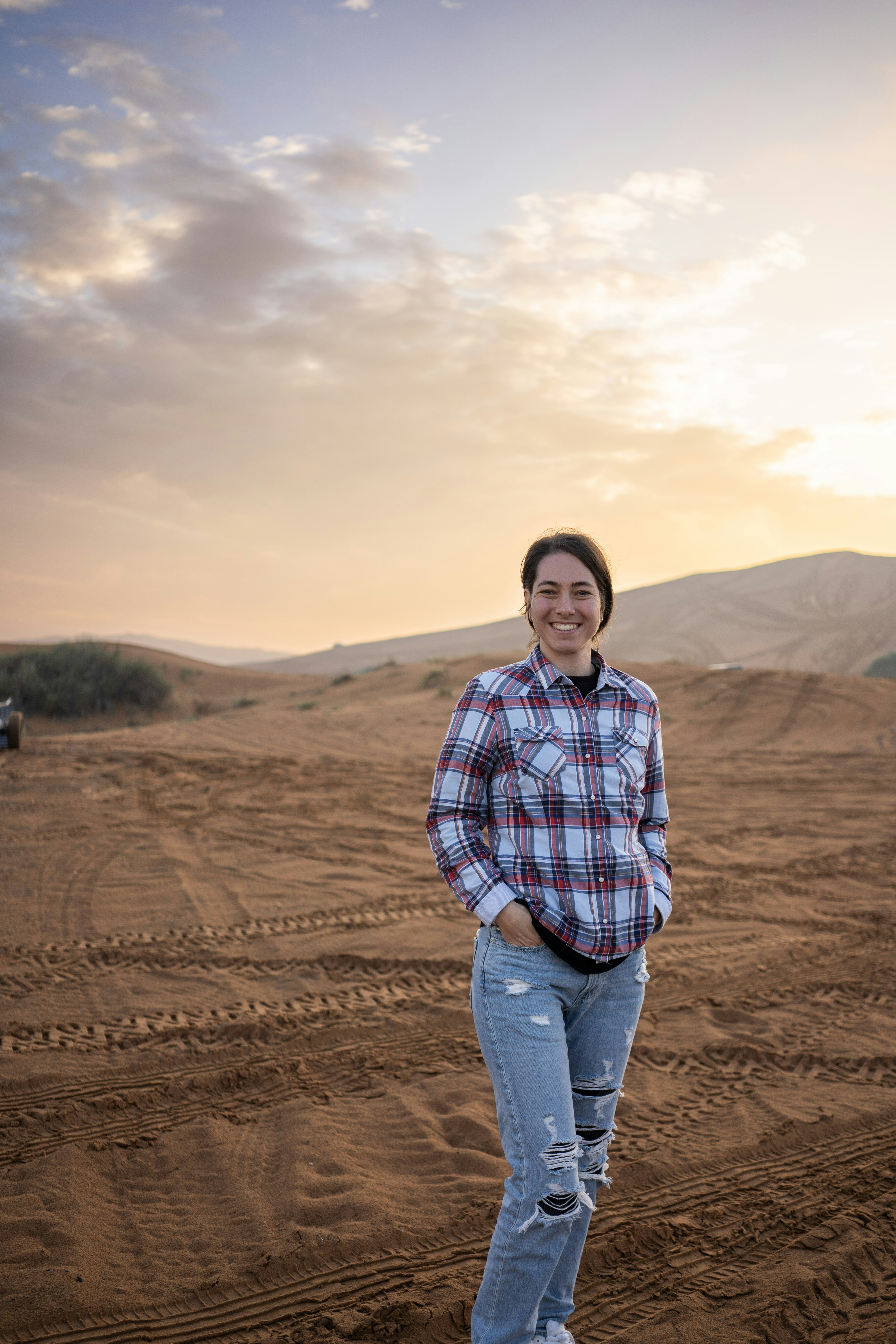 A smiling woman stands confidently in the desert at sunset, wearing a plaid shirt and distressed jeans. The serene landscape features rolling sand dunes and a colorful sky.