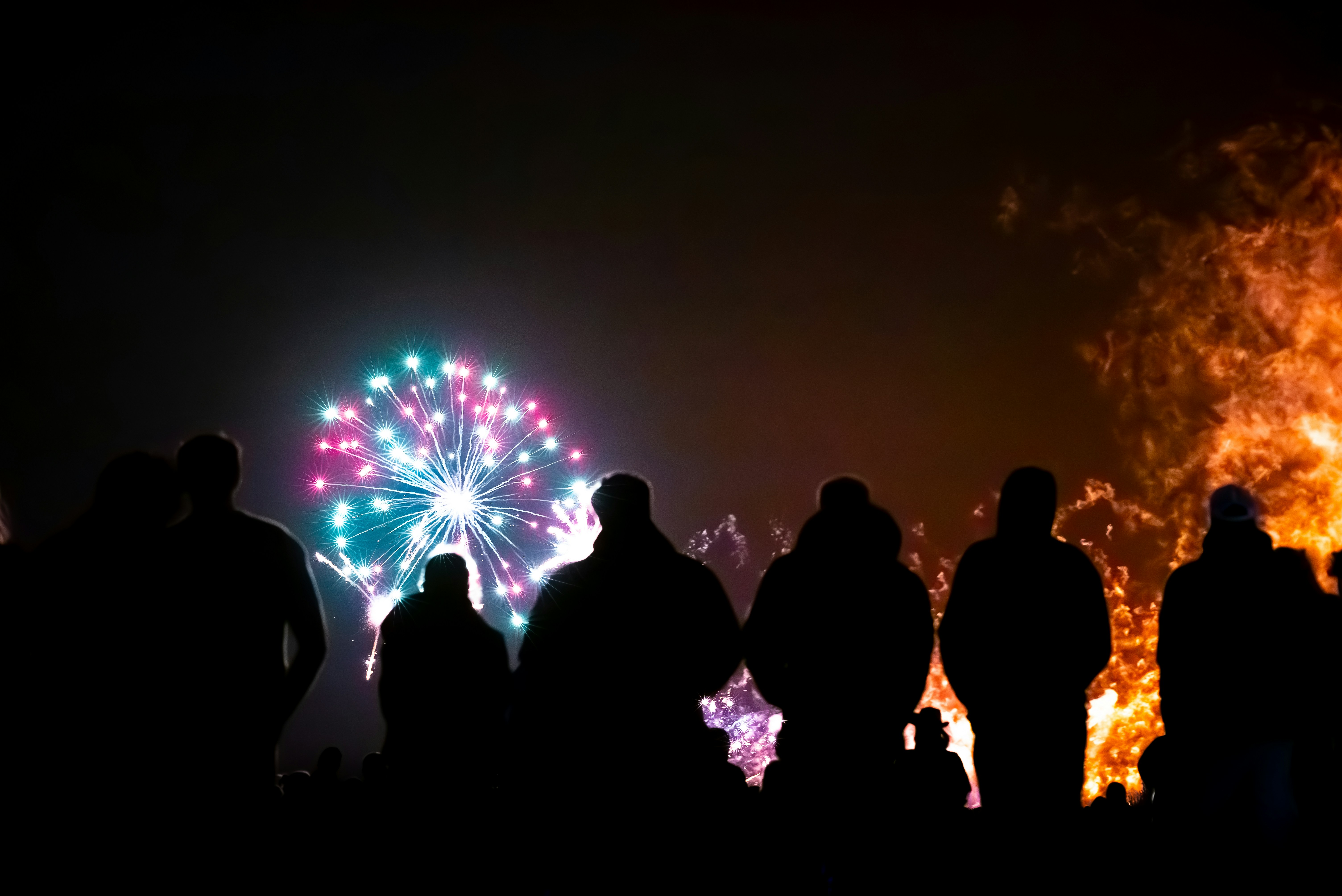 A crowd watches as vibrant fireworks bloom in the distance, their silhouettes glowing softly against the fiery orange haze of the night.