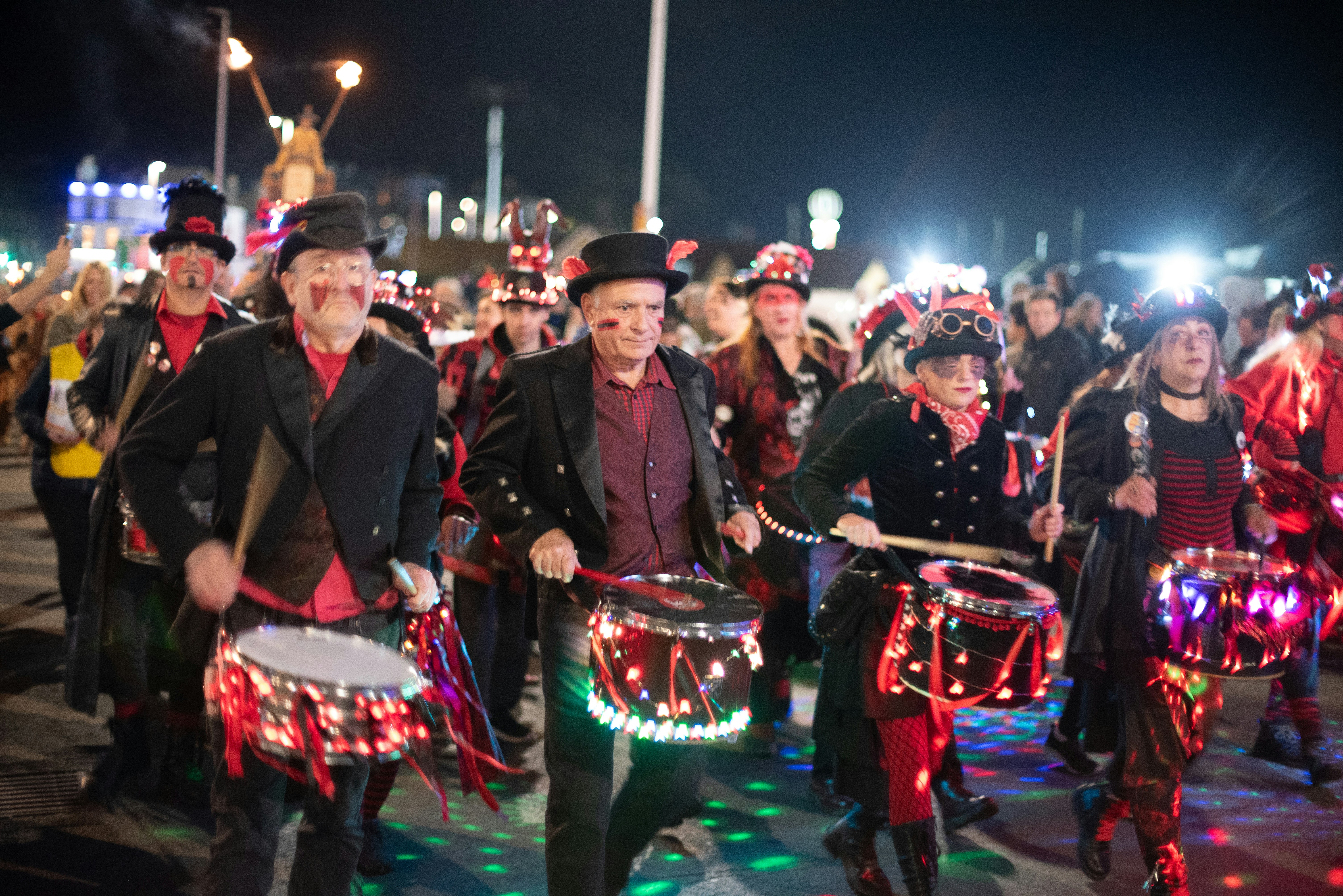 A vibrant group of drummers dressed in red and black march through the night, their illuminated drums beating in rhythm with the festival’s electric energy. | People in costumes playing drums in a nighttime parade.