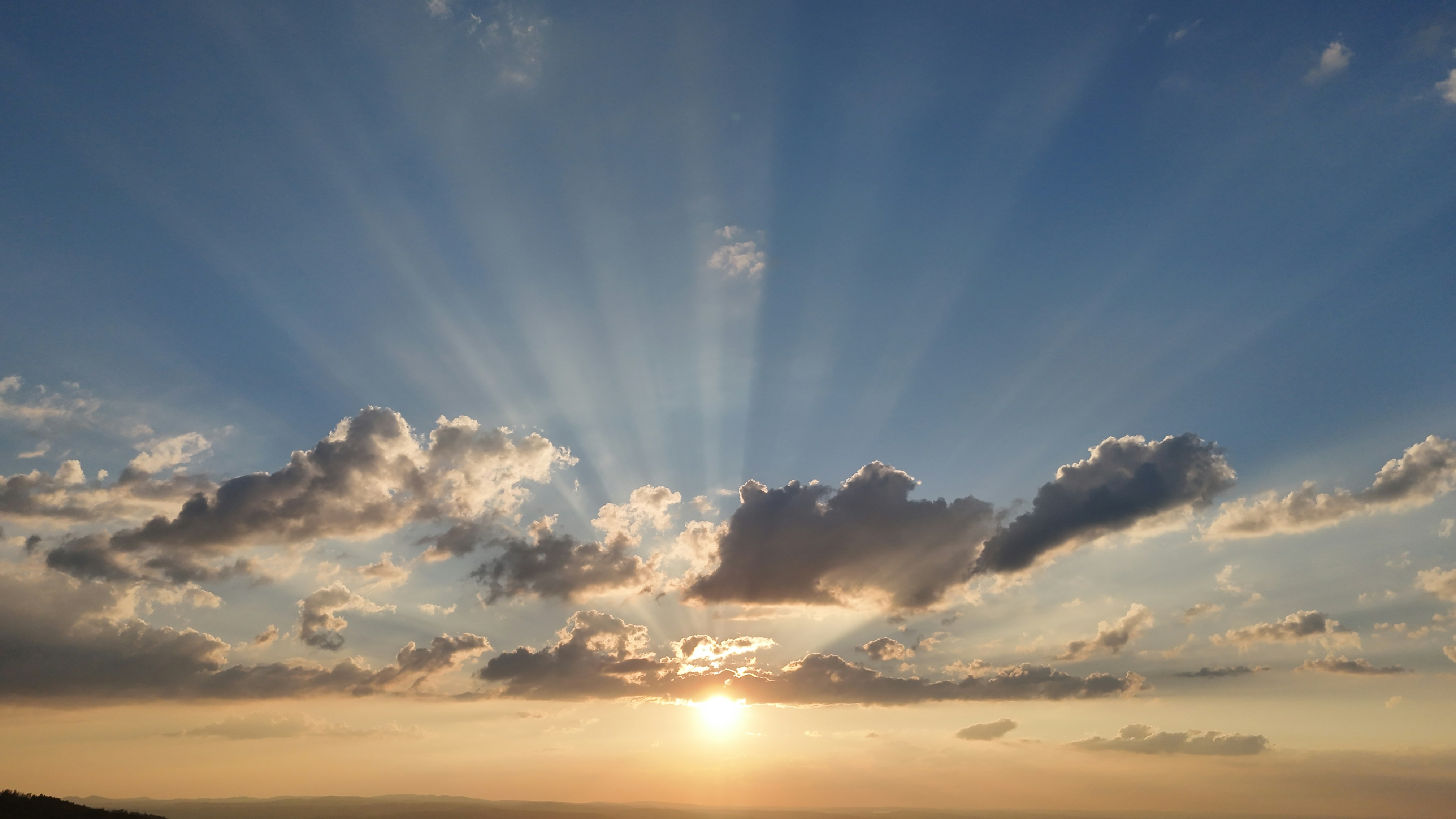 Golden sun setting behind a horizon, rays of light streaming through scattered clouds, creating a dramatic sky.