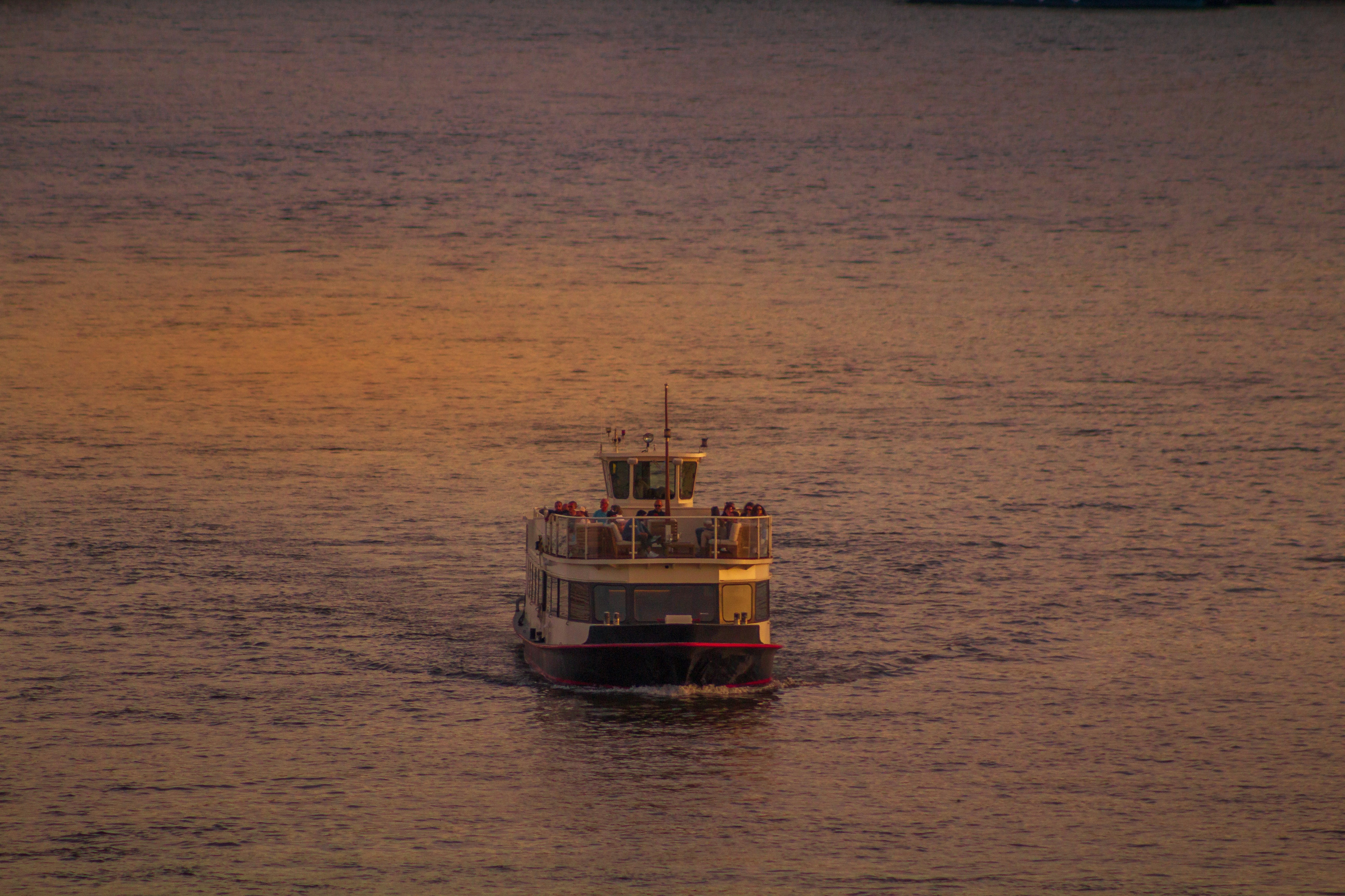A boat gliding through calm waters at sunset, reflecting hues of orange and purple. The passengers enjoy a tranquil journey amidst the serene backdrop.