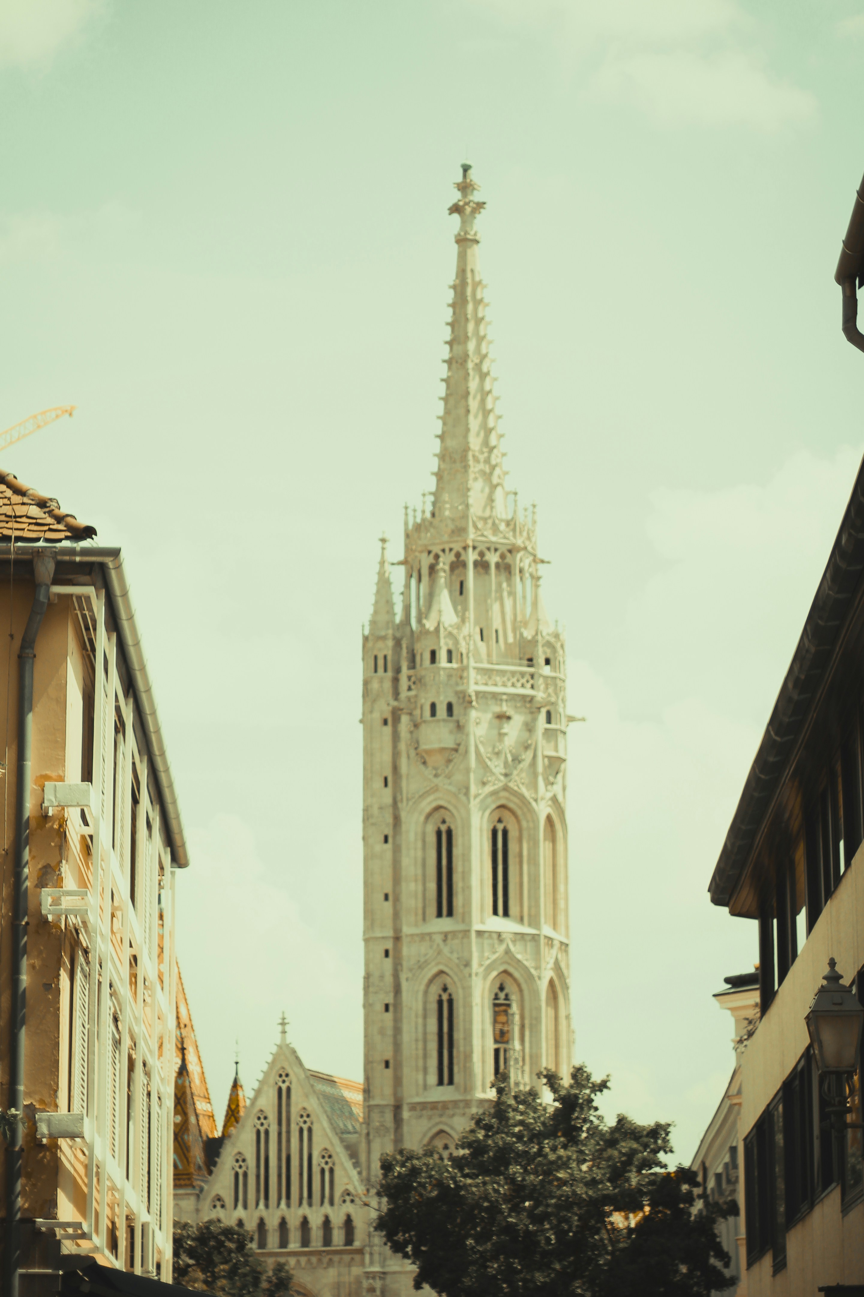 A majestic Gothic tower rises above the narrow streets, framed by historic buildings and lush greenery.