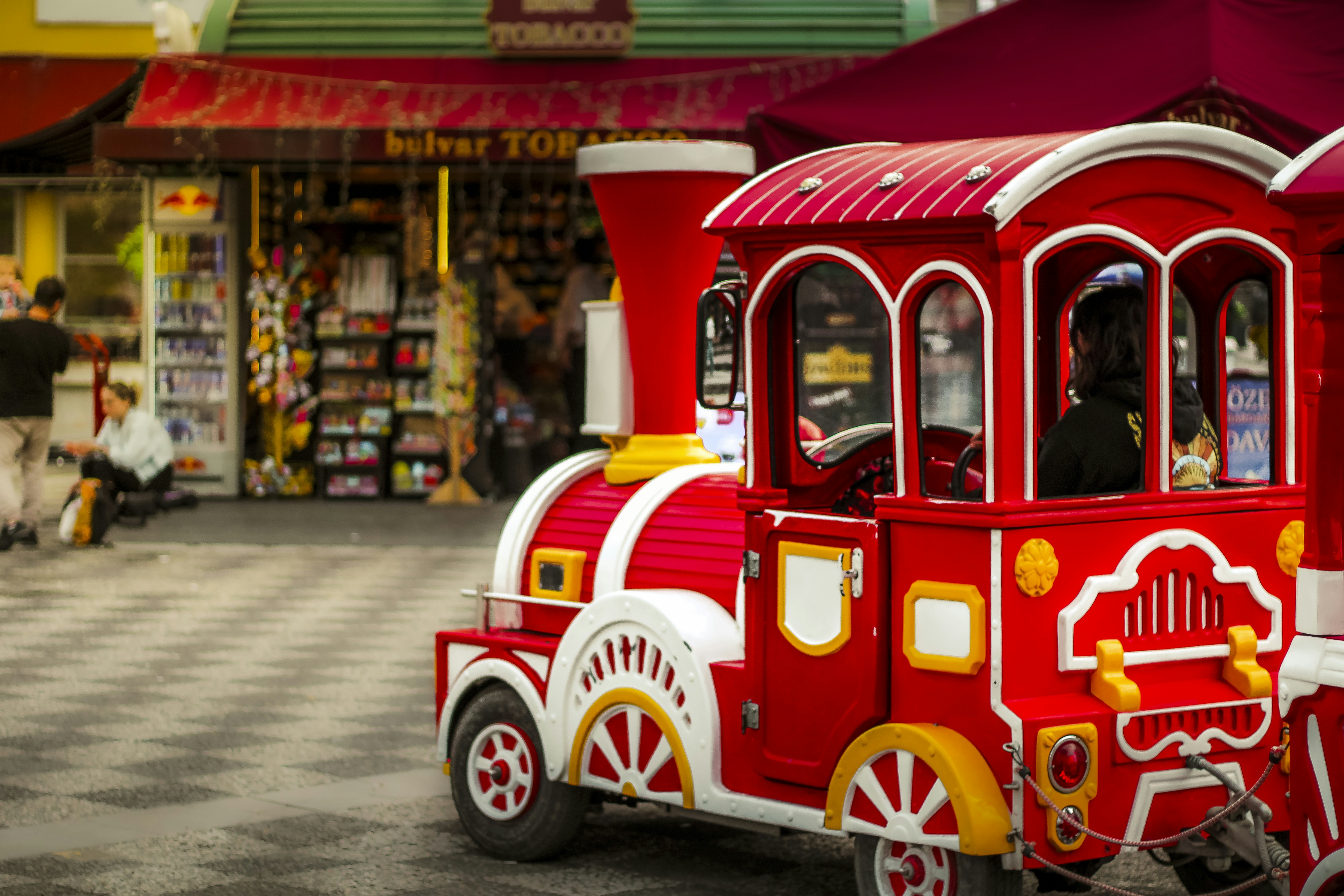 Colorful miniature train parked in a lively market, surrounded by vibrant shops and visitors. The scene captures the essence of fun and exploration.