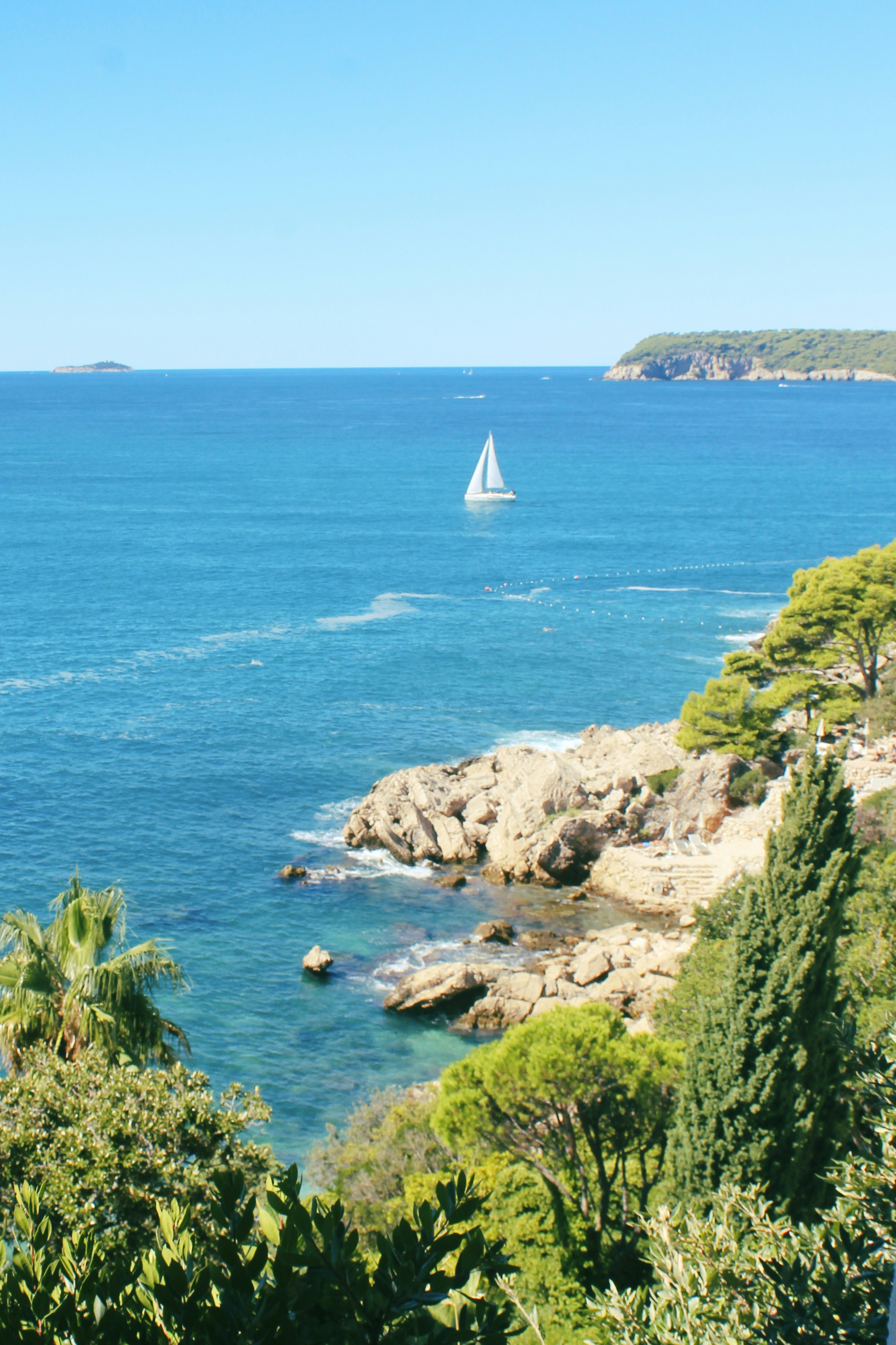 Sailboat on a bright blue ocean near rocky shore.