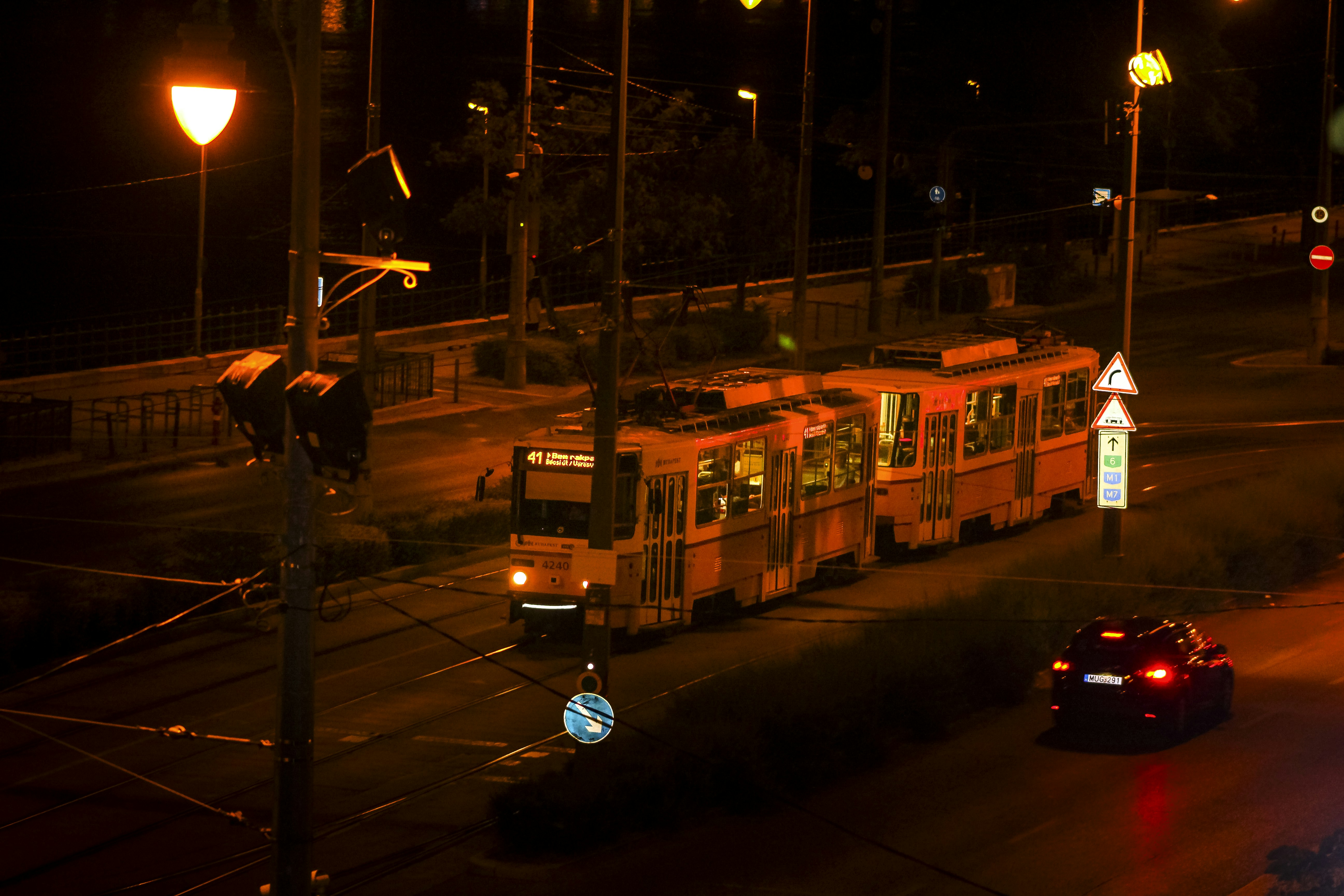 Tram navigating through a quiet city street illuminated by streetlights at night. Traffic signs and a parked car add to the urban ambiance.