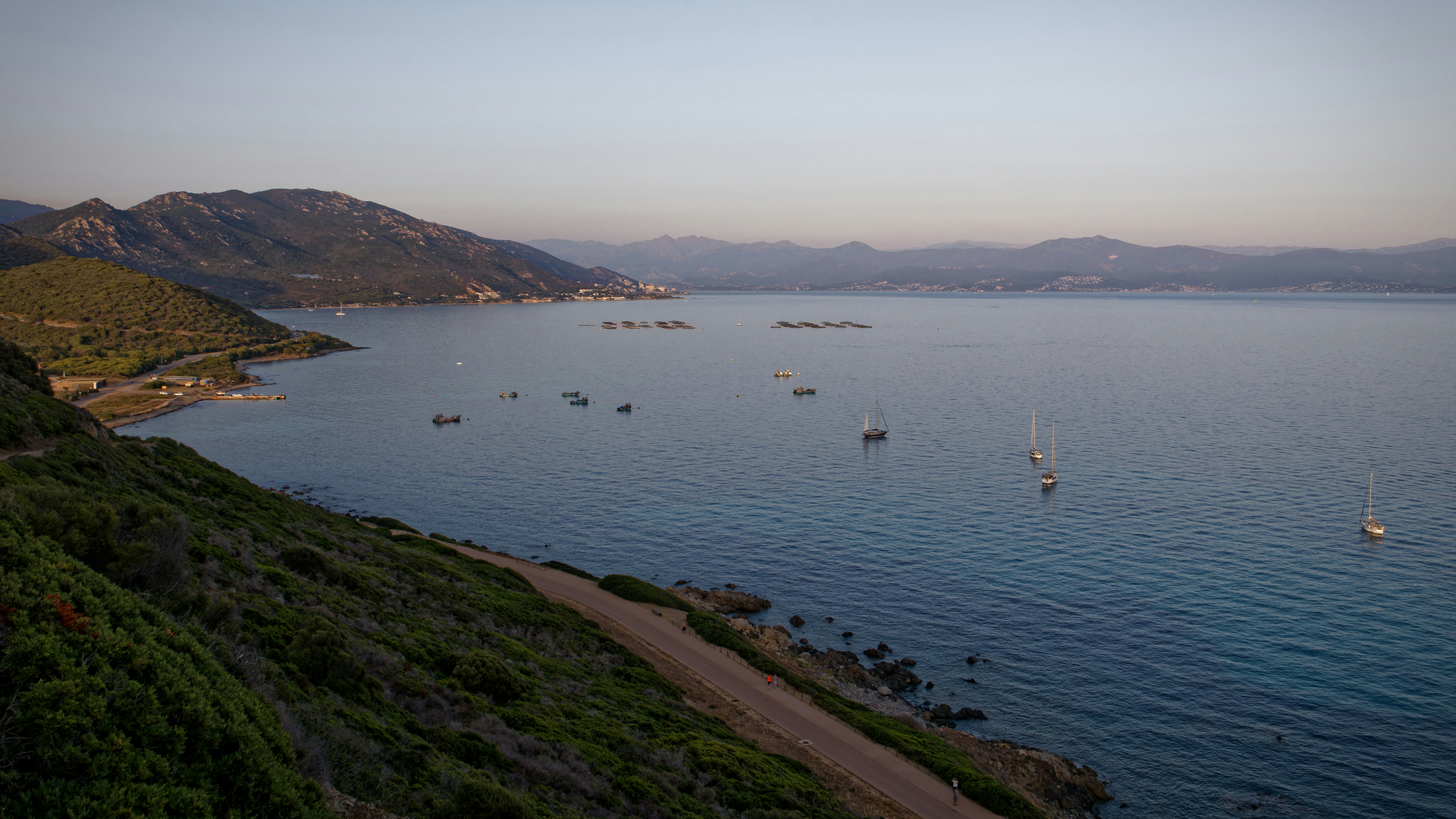 Sailboats dot a calm bay near a green, hilly coastline.
