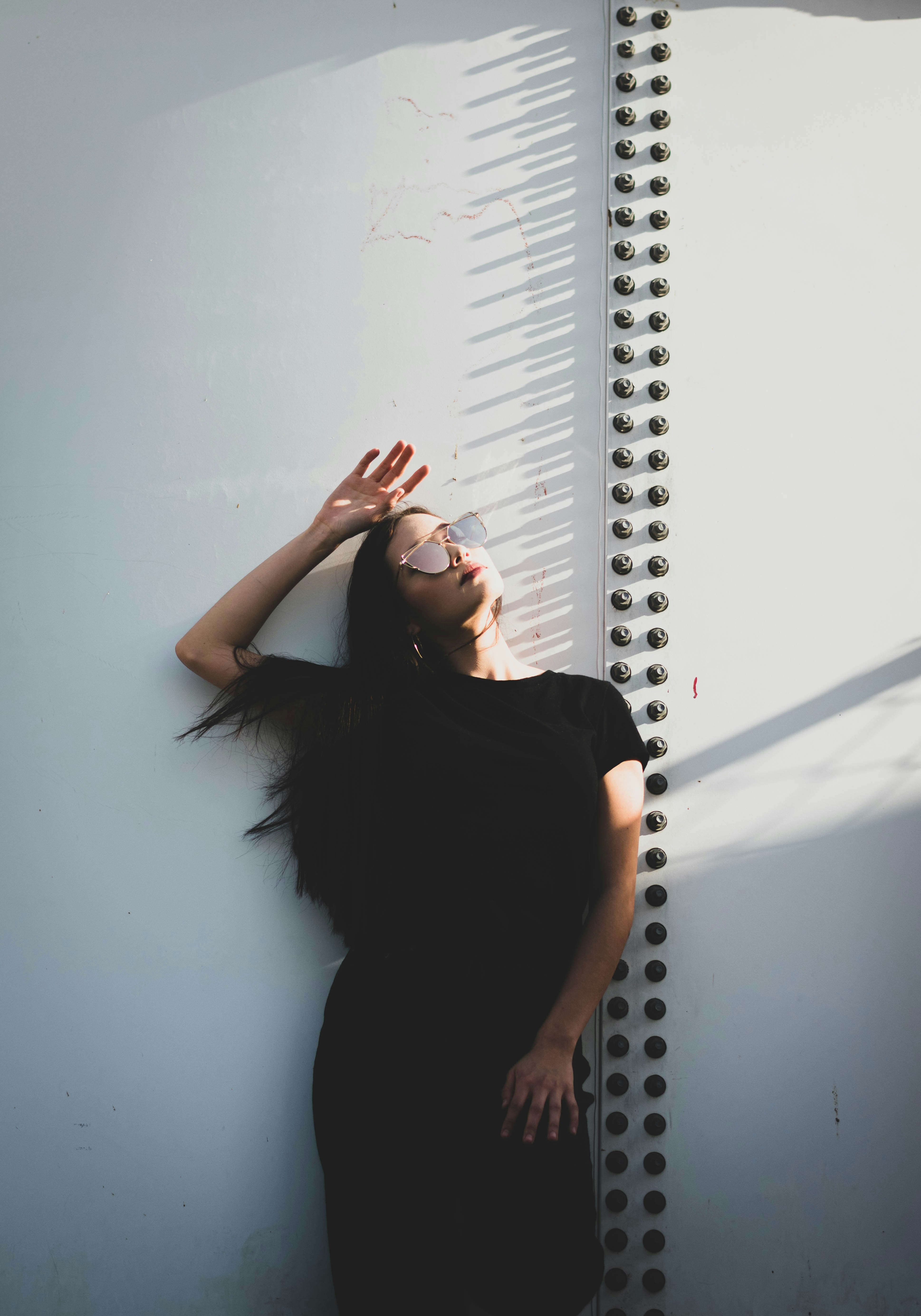 A woman poses against a white wall, her hand raised to her forehead, casting intriguing shadows. The interplay of light and texture adds depth to the scene.