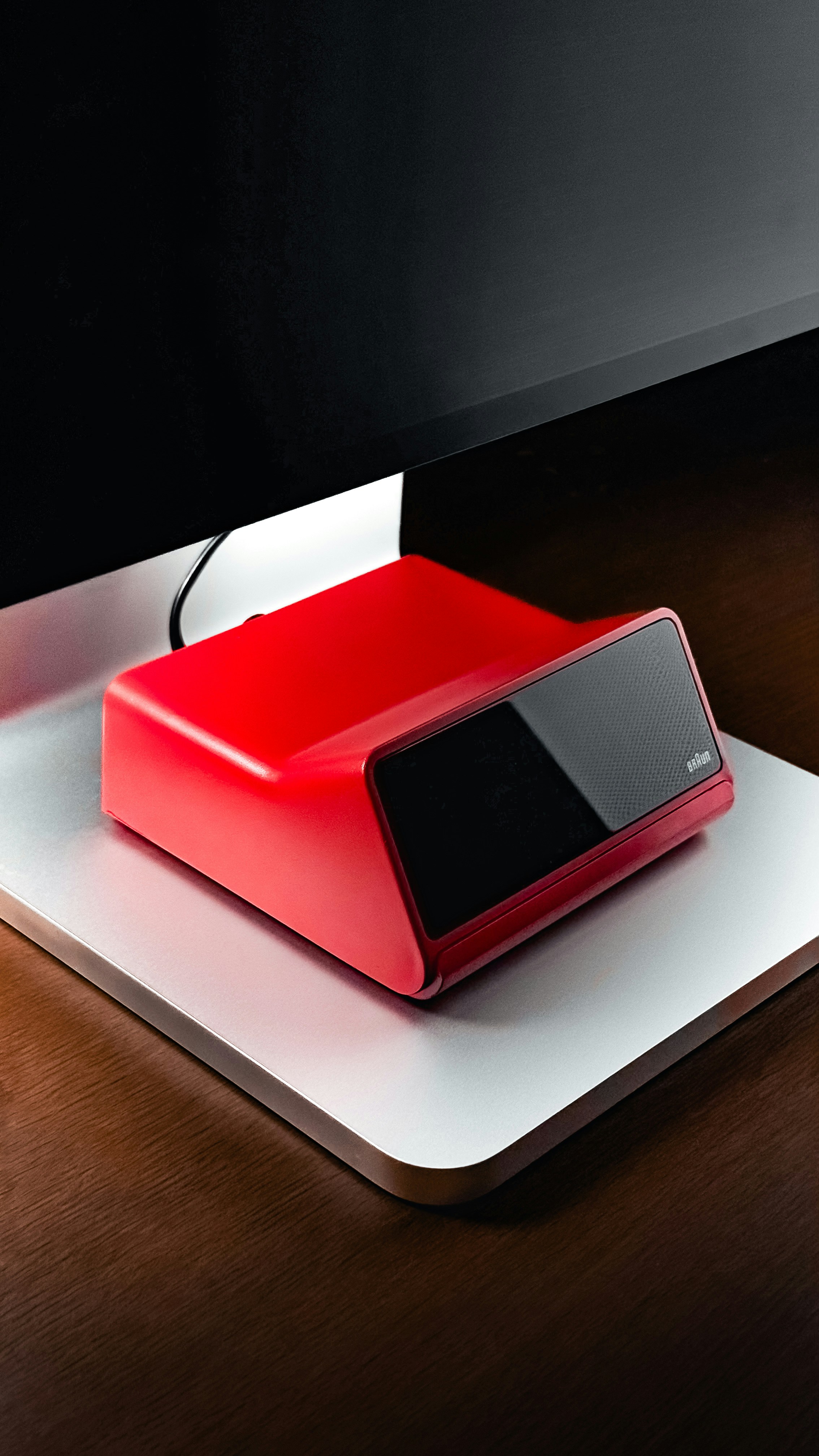 A detailed close-up of the iconic Braun DN 40 clock designed by Dieter Rams, featuring its striking red body and minimalist black display. The composition highlights Rams’ “less but better” philosophy, blending functionalism with timeless modern design aesthetics. | Red scanner device in front of computer monitor