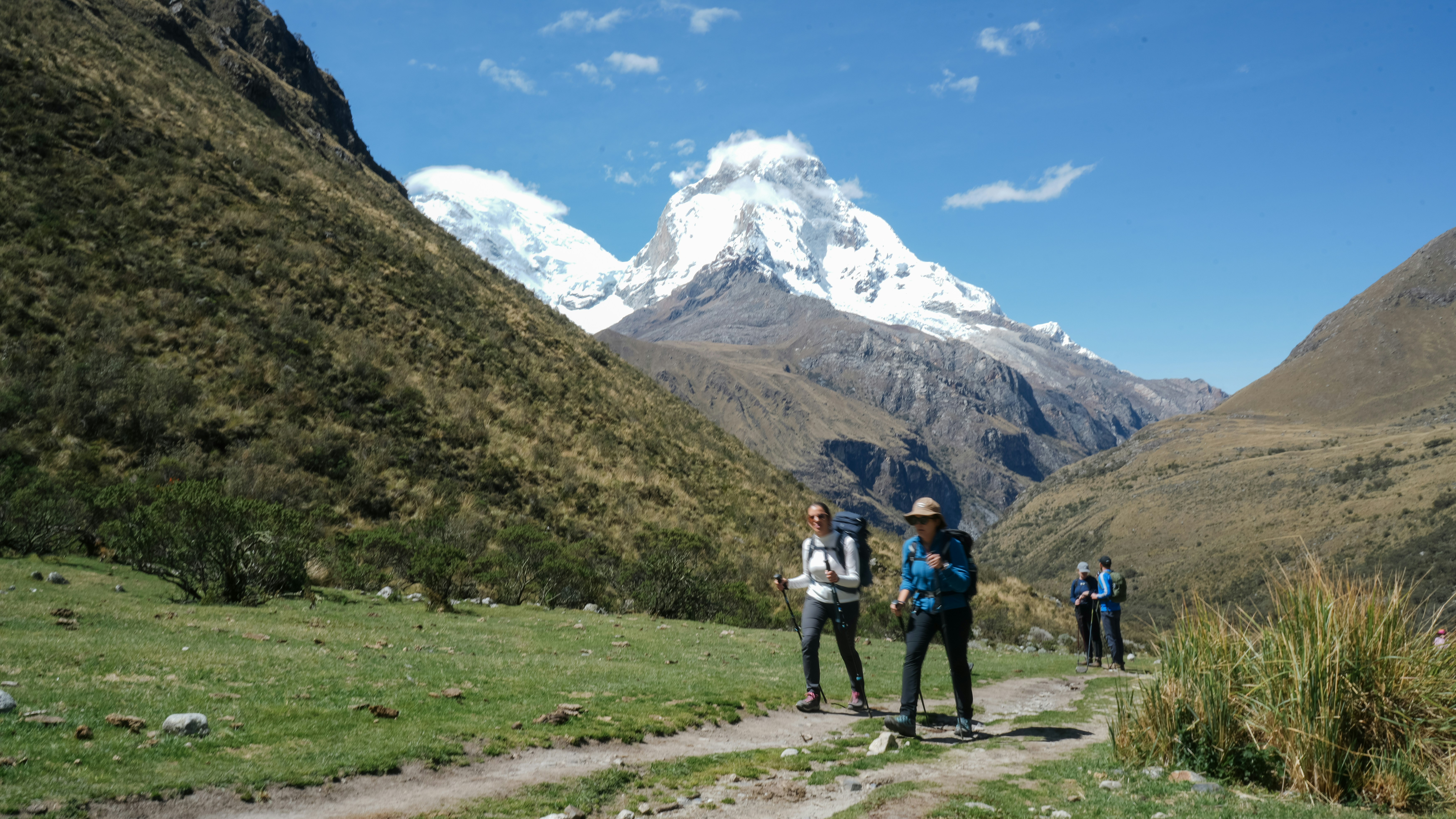 Hikers walk a mountain trail toward snow-capped peaks.