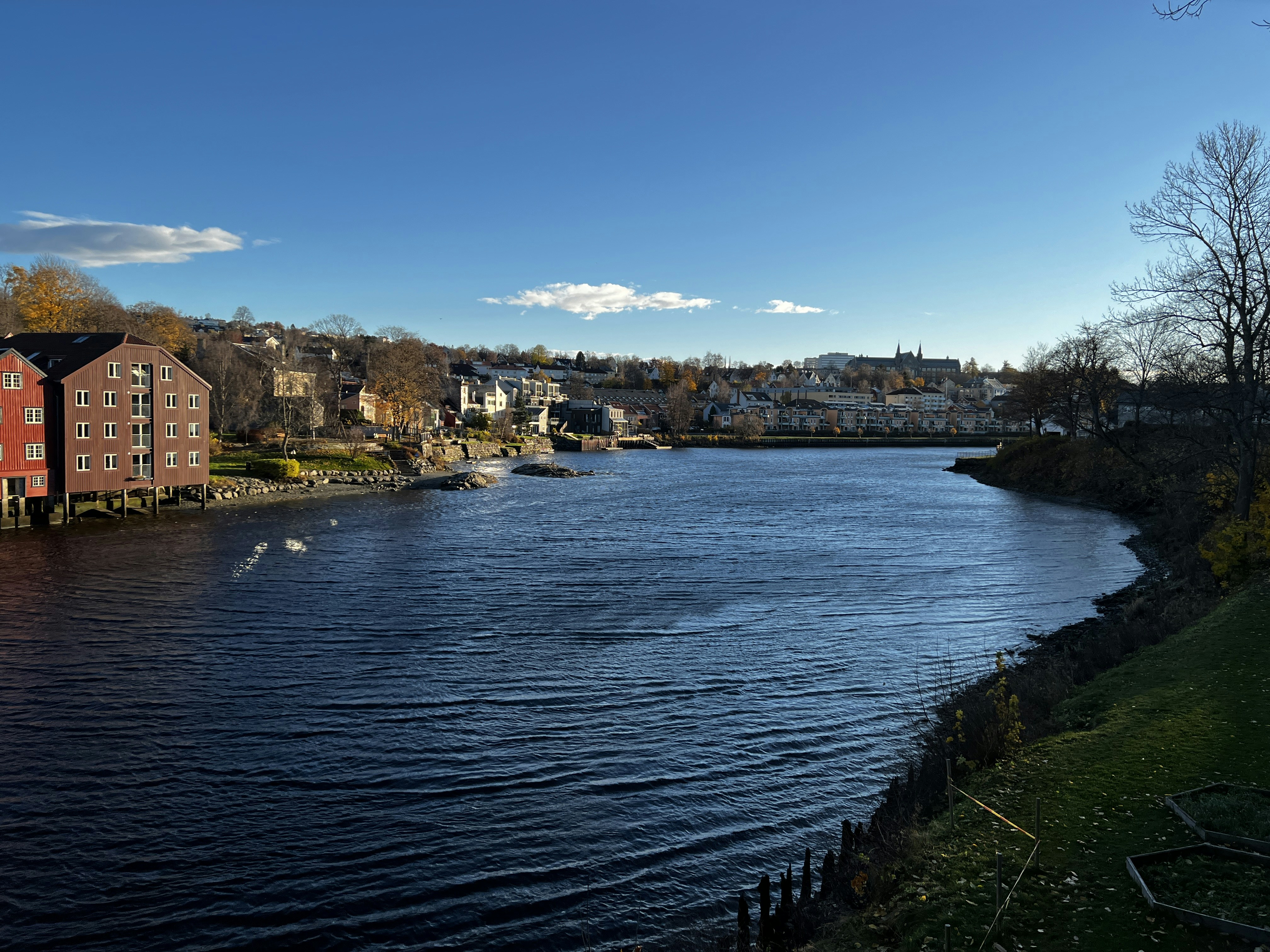 Tranquil waters flow beneath the charming houses of Kjøpmannsgata, with a majestic bridge connecting the banks in the picturesque city of Trondheim. | River flowing through a town with buildings and trees.