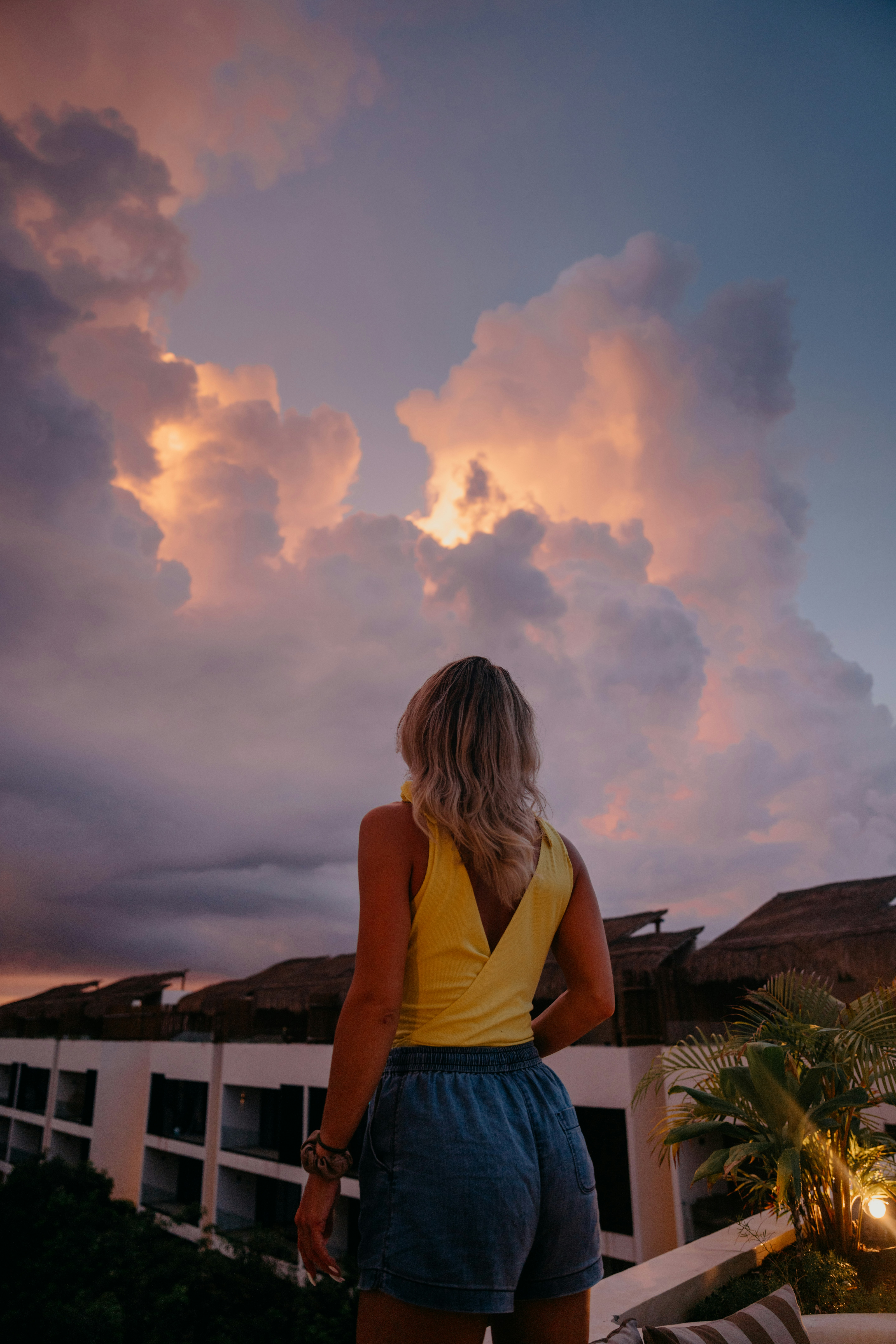 A woman stands on a balcony, gazing at the colorful clouds illuminated by sunset hues. The scene conveys a serene pause amidst a vibrant sky.