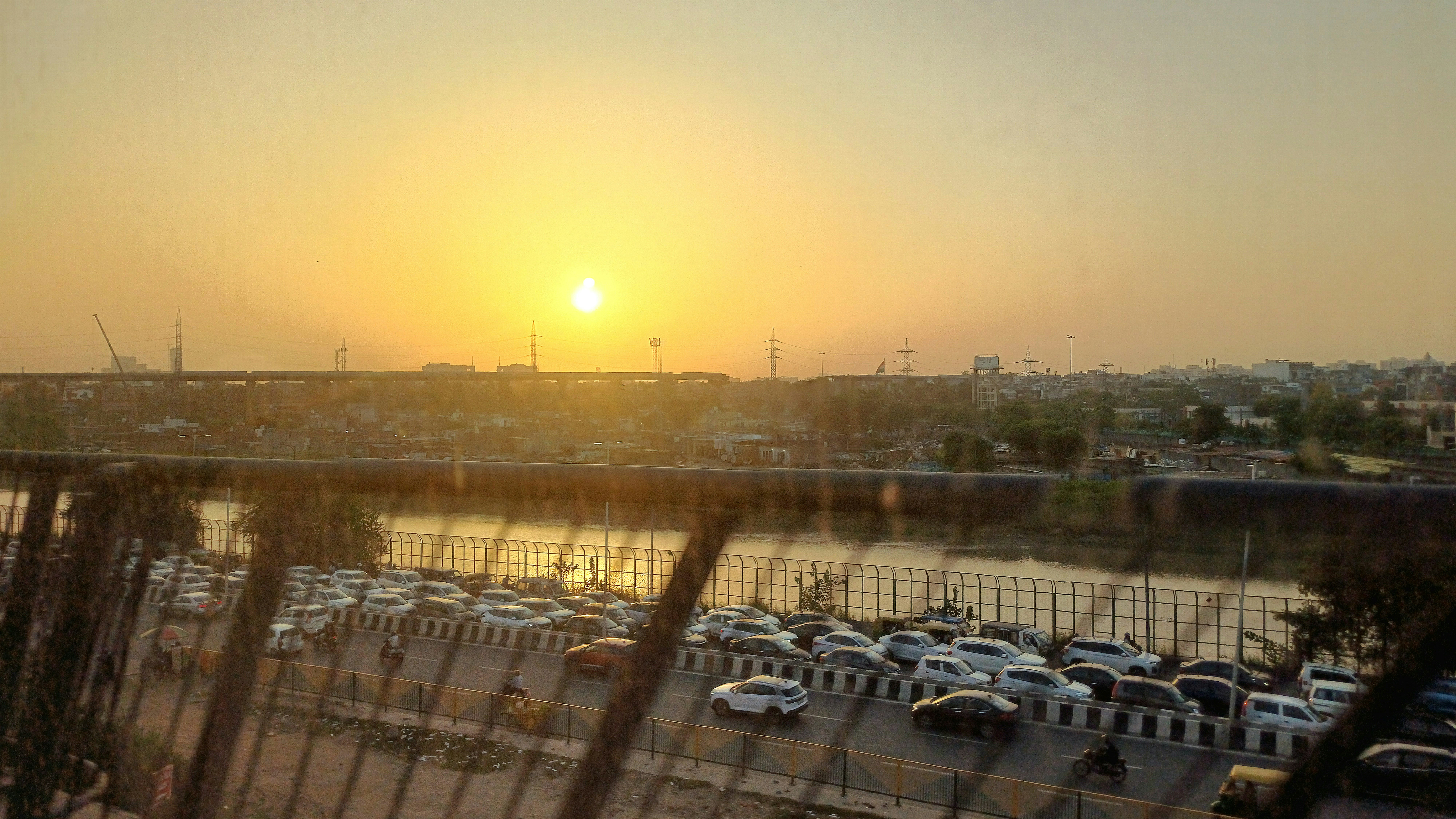 Sunset casting a warm glow over a bustling cityscape, with cars lined along the riverbank and silhouettes of buildings in the background.
