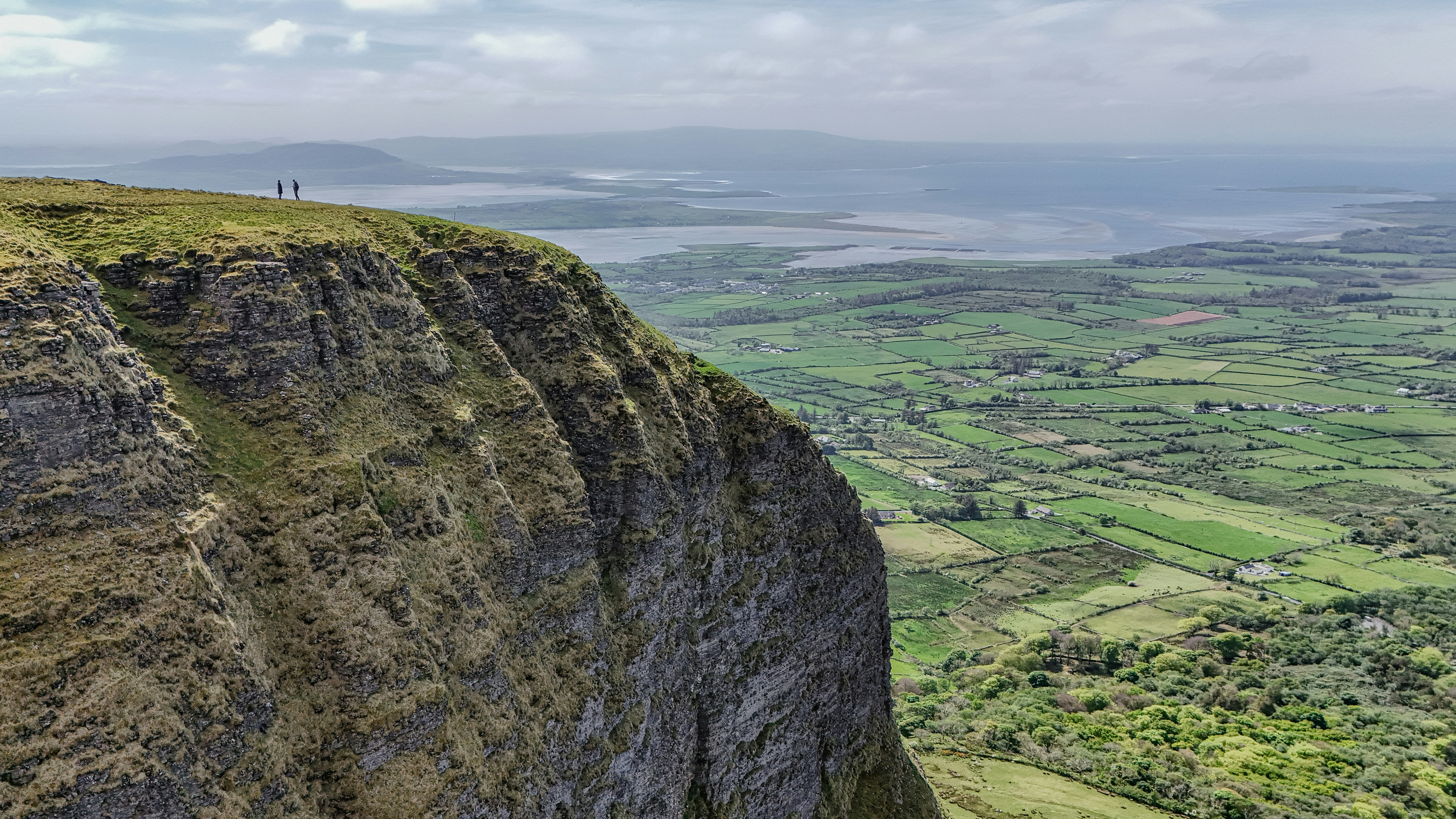 Two hikers stand on the edge of Benbulben, taking in the view. In the foreground, the mountain’s rugged texture contrasts with the vast landscape of green fields and water stretching out below. | Two people stand on a cliff overlooking a valley.
