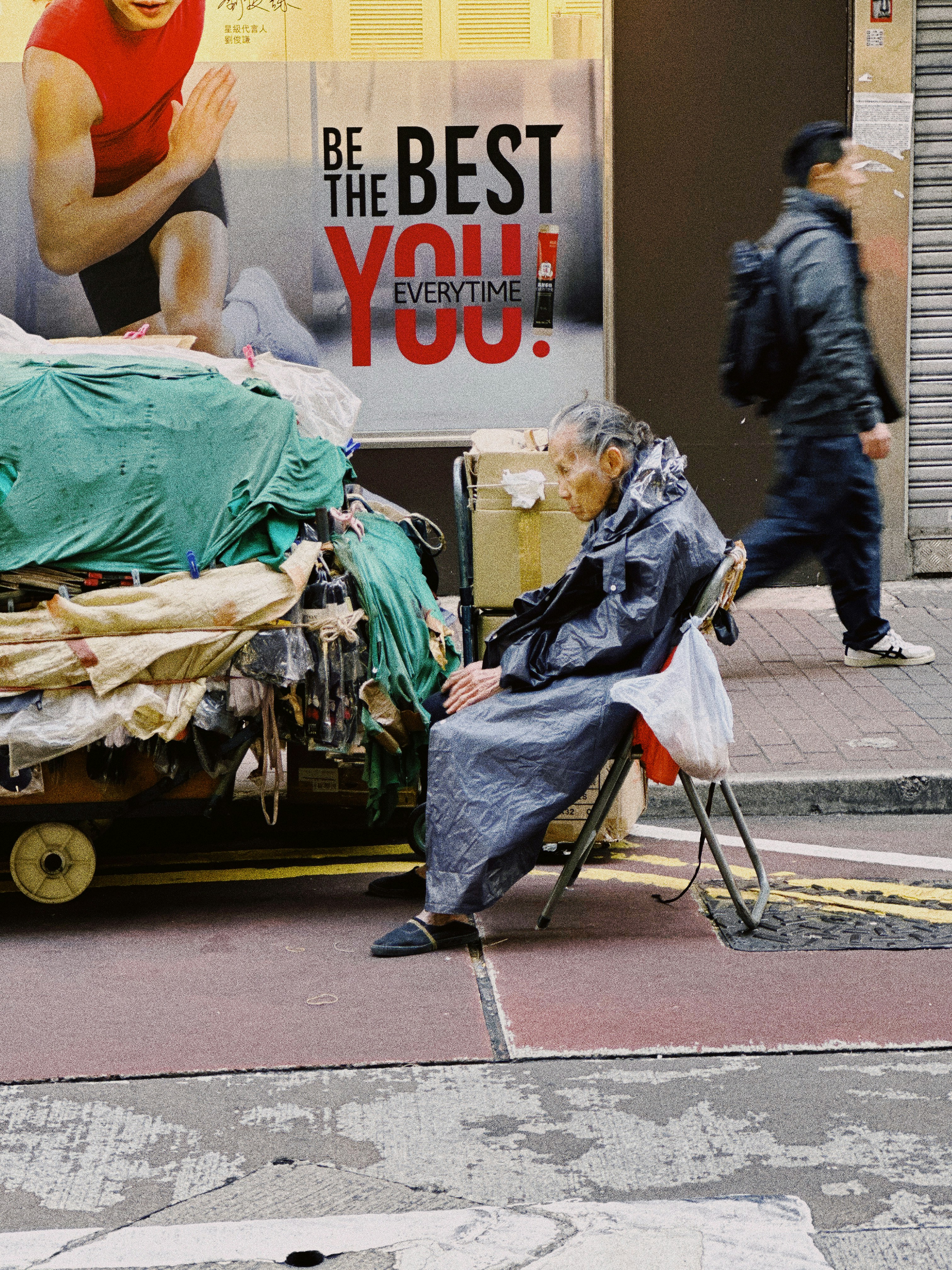 Elderly person sits by cart on city street.
