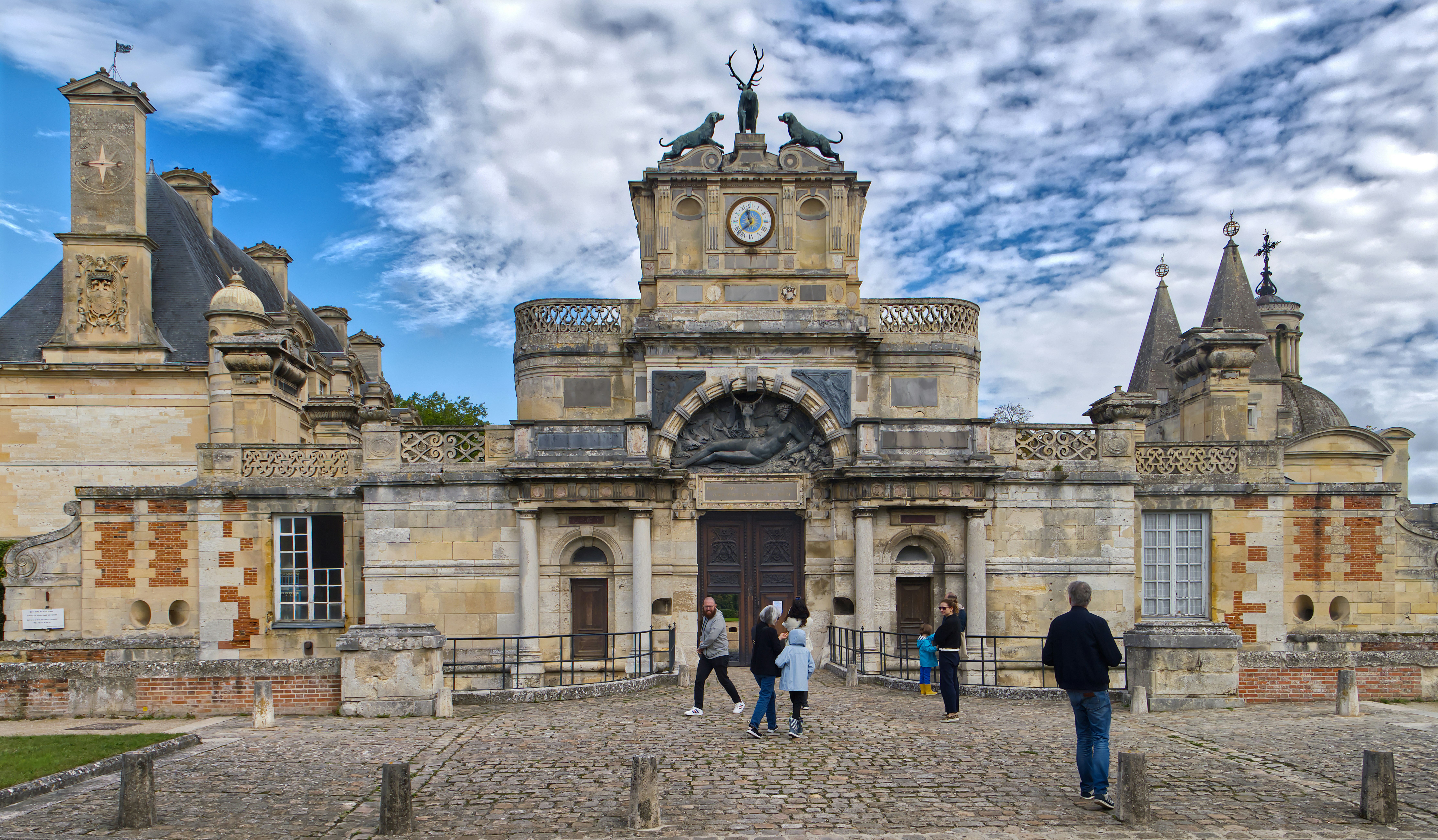 J’ai pris cette photo juste avant la visite du château d’Anet, un site emblématique de la Renaissance française, édifié au XVIᵉ siècle pour Diane de Poitiers, la favorite du roi de France Henri II. Depuis l’esplanade, la façade d’entrée impressionne par son équilibre et la richesse de ses détails sculptés. On y distingue l’horloge centrale, surmontée de deux chiens et d’un cerf, symboles de la chasse et de la déesse Diane. | People walking towards ornate stone building entrance under cloudy sky.