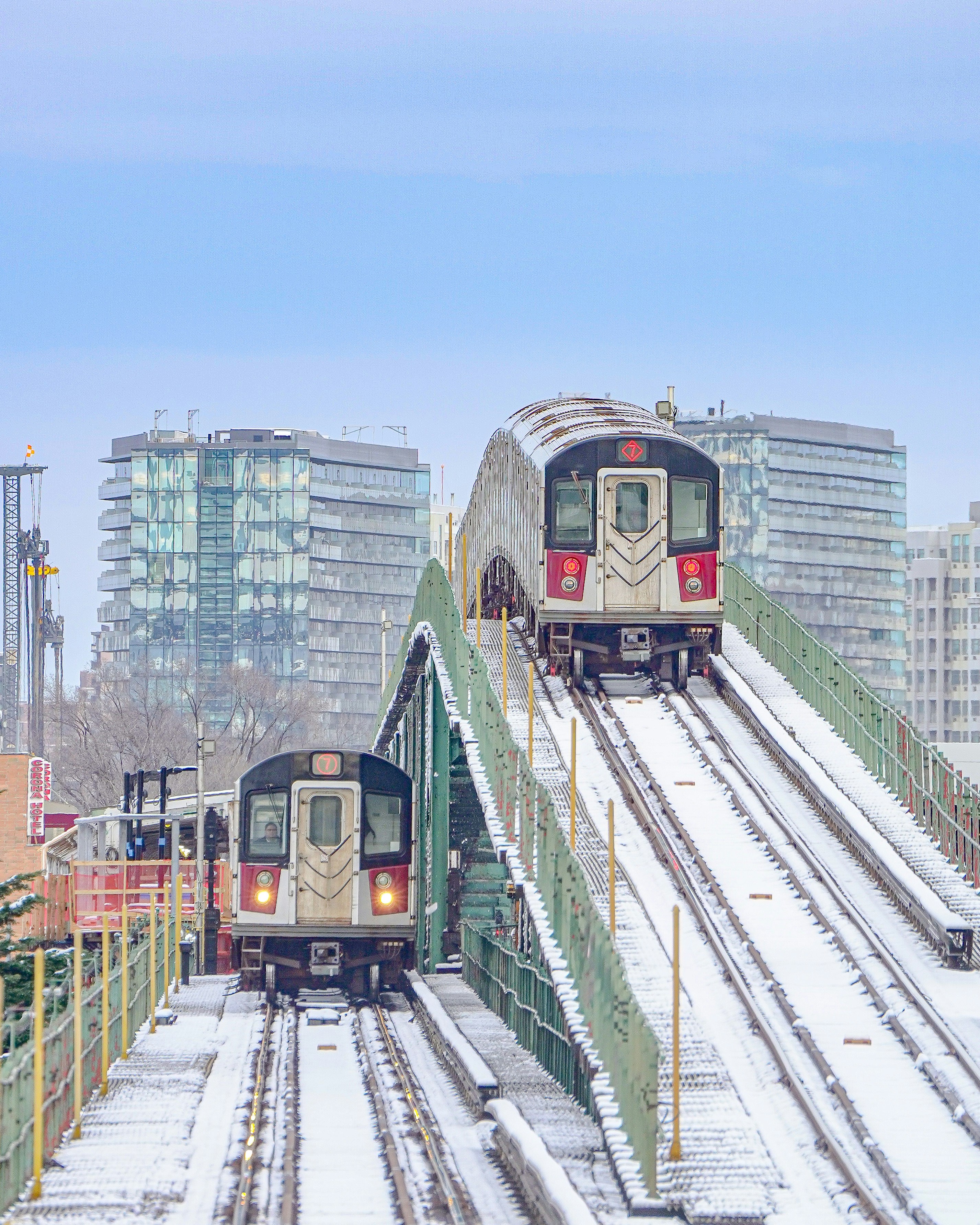 Two subway trains navigating a snowy elevated track, framed by modern buildings in the background.