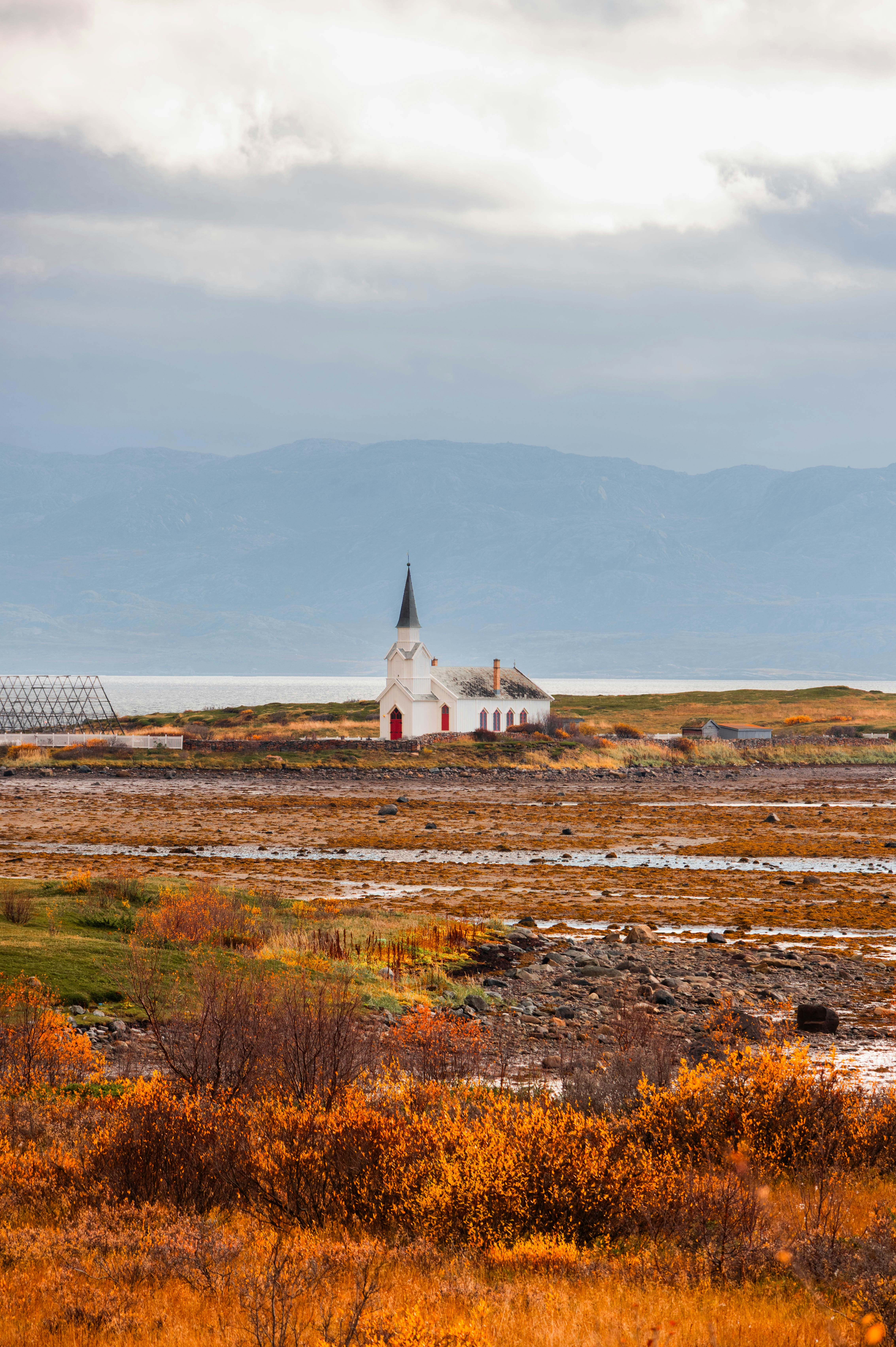 A quaint white church with a tall spire stands alone amidst a serene landscape of autumn foliage and distant mountains. The scene captures the essence of solitude and tranquility.