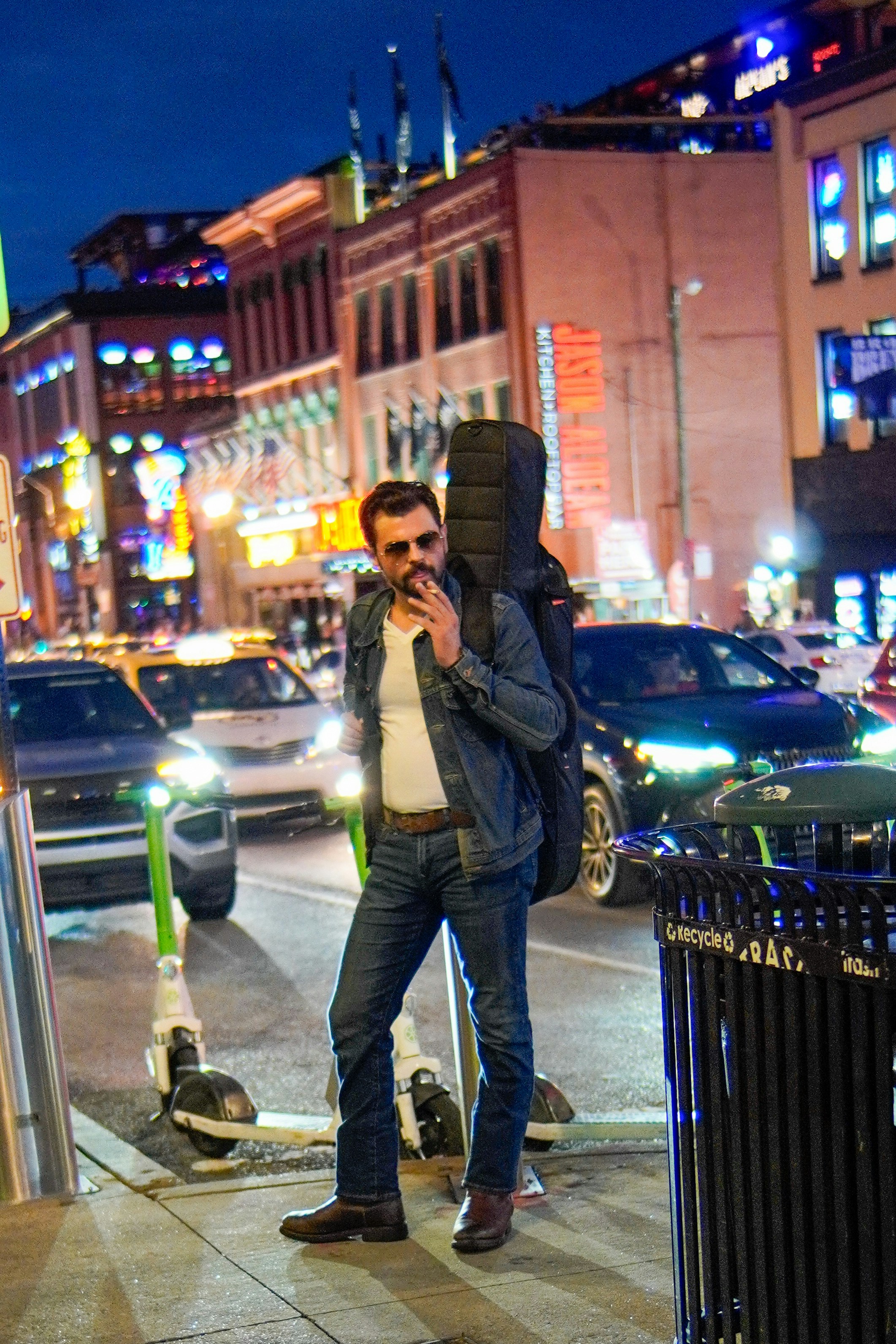 Man with a guitar stands on a bustling street, surrounded by vibrant nightlife and traffic. Neon lights illuminate the scene.