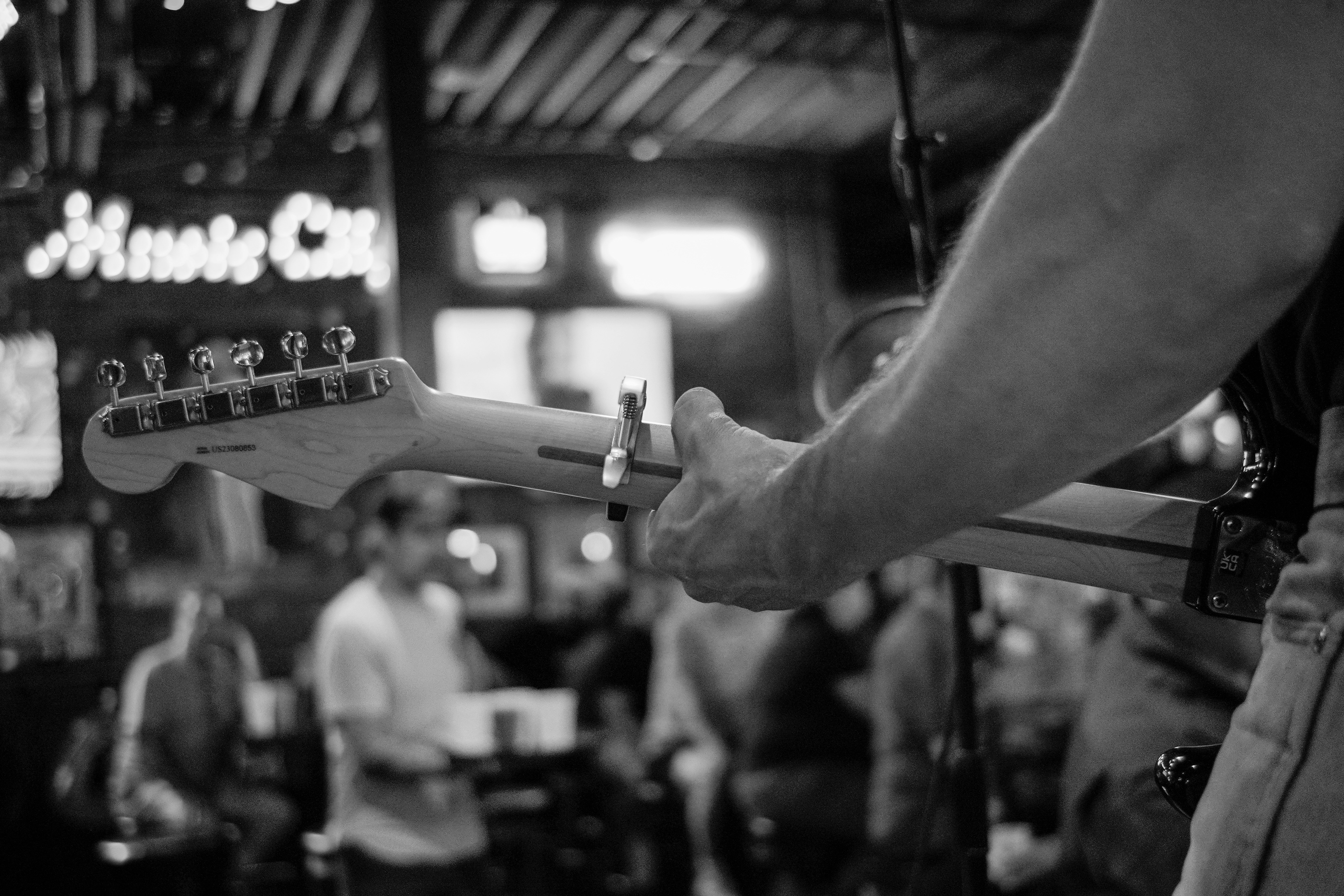 Close-up of a guitarist's hand gripping the neck of an electric guitar, with a lively bar scene blurred in the background.