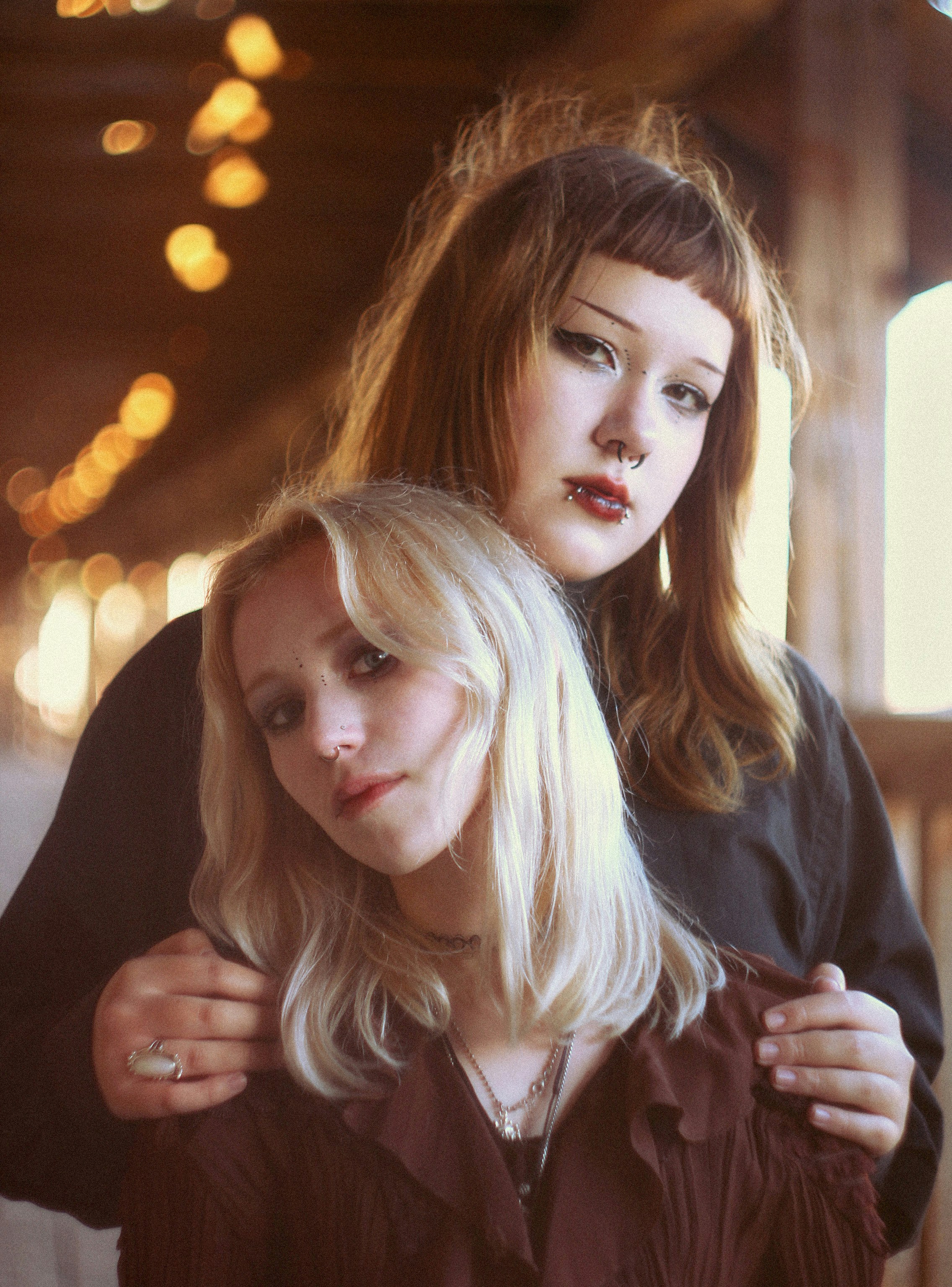 Two young women share an intimate moment, showcasing their unique styles and expressions against a softly blurred background of warm lights.