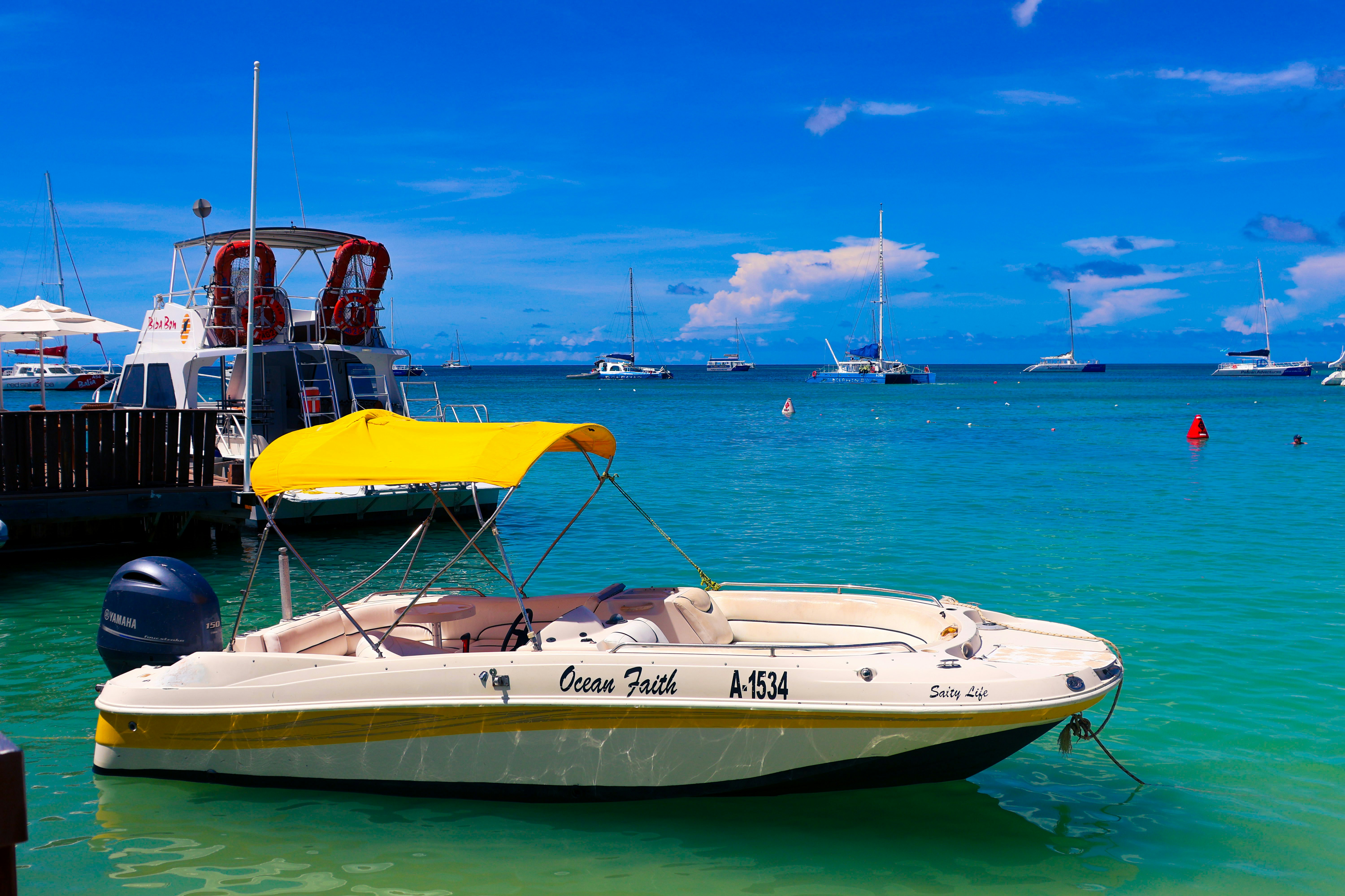 A white motorboat with a yellow canopy anchored in turquoise waters, surrounded by other vessels and a clear blue sky.