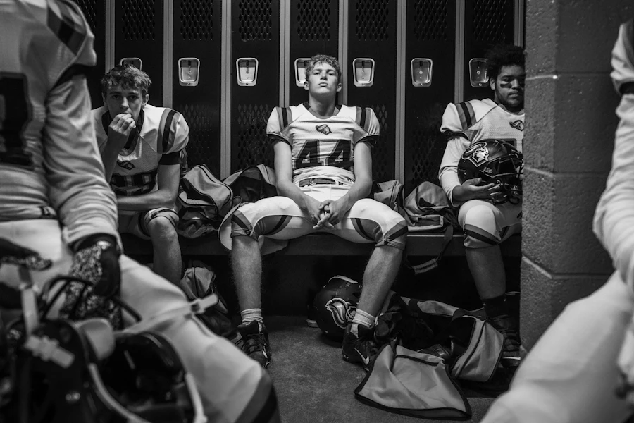 High-school football player wearing number 44 seated between teammates in a locker room, eyes closed and hands clasped.