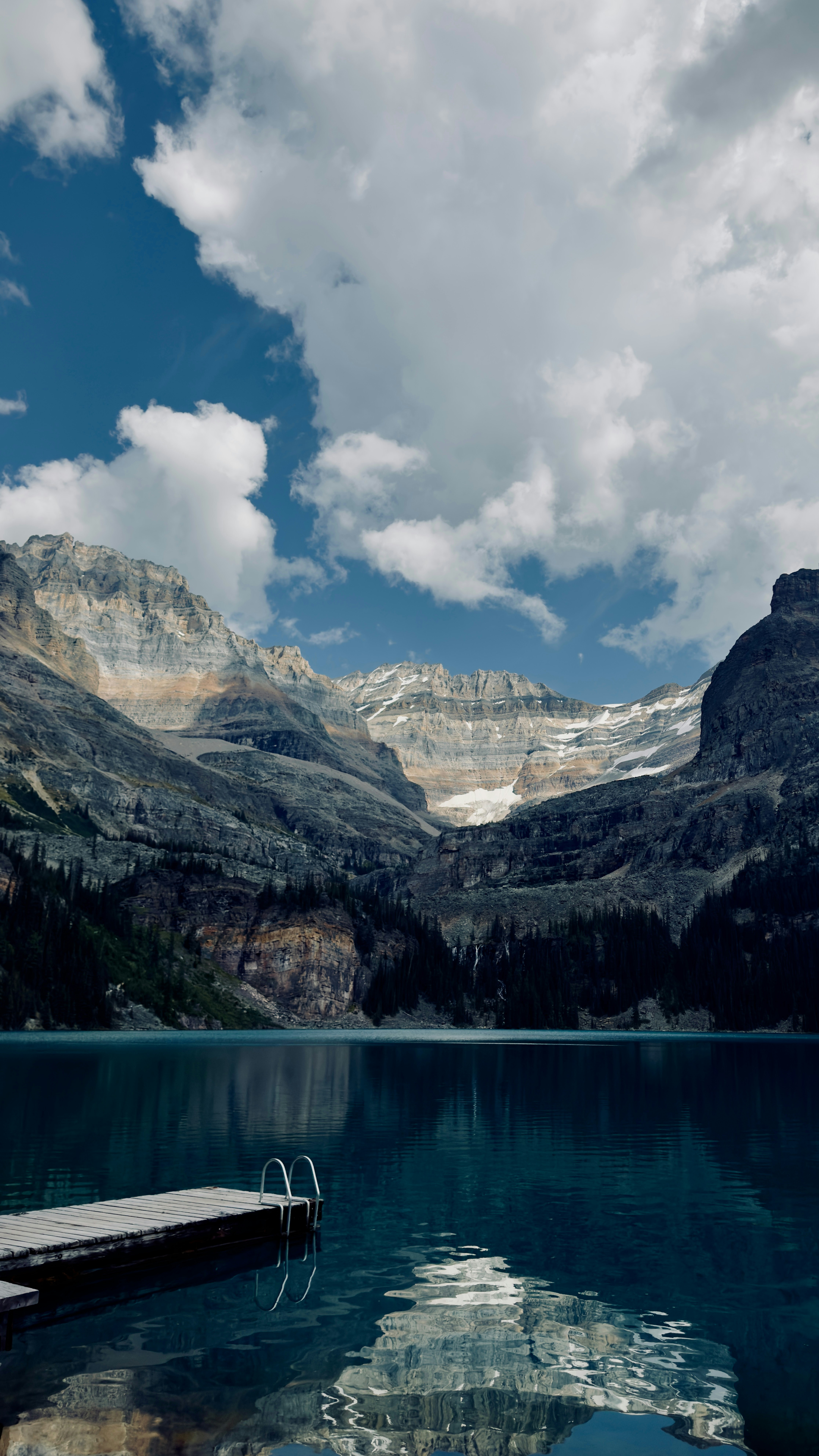 Serene mountain lake reflecting towering peaks and a dramatic sky, with a wooden dock in the foreground.