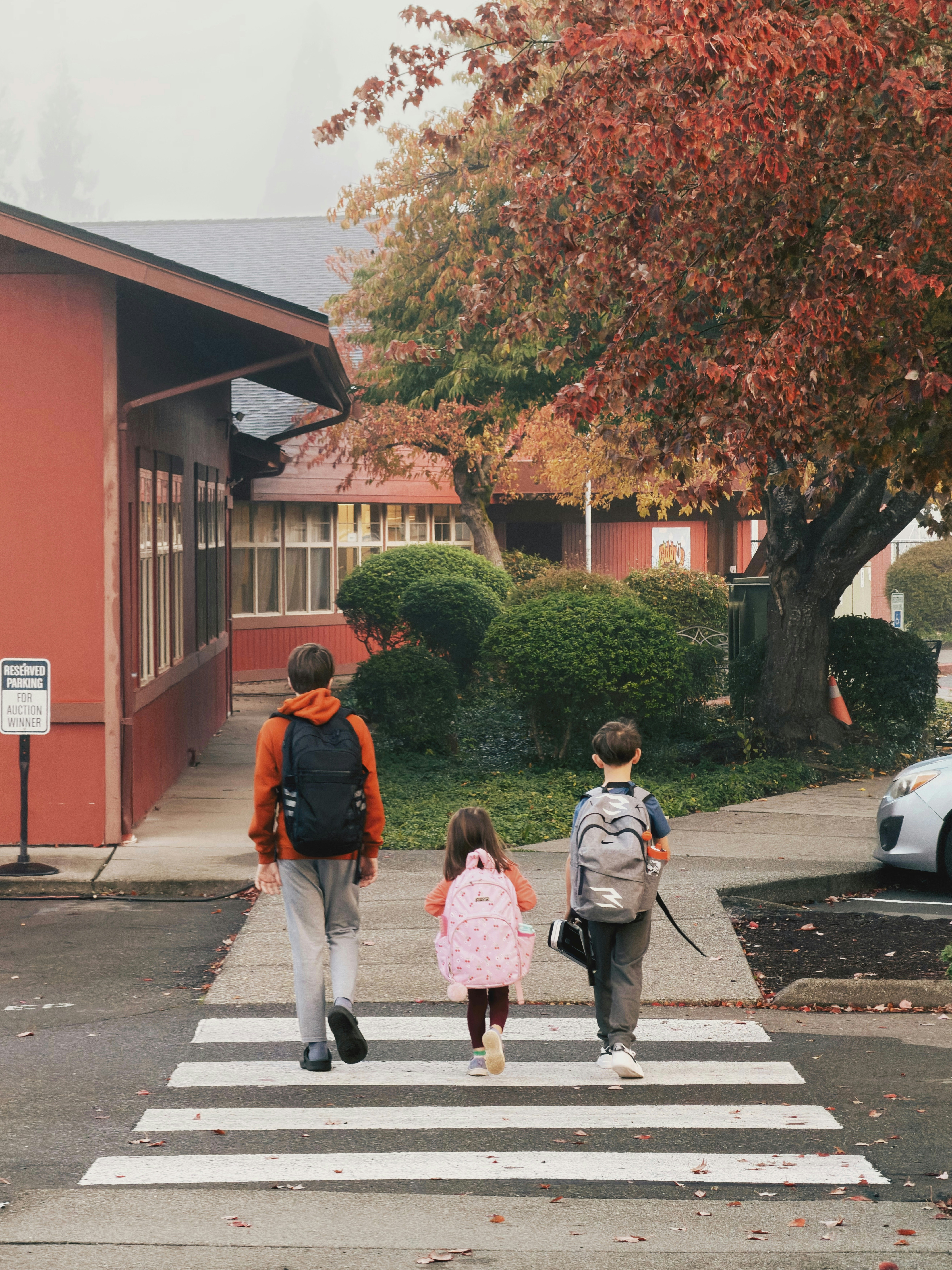 Three children crossing a street adorned with vibrant autumn leaves, leading towards a school building in the background.