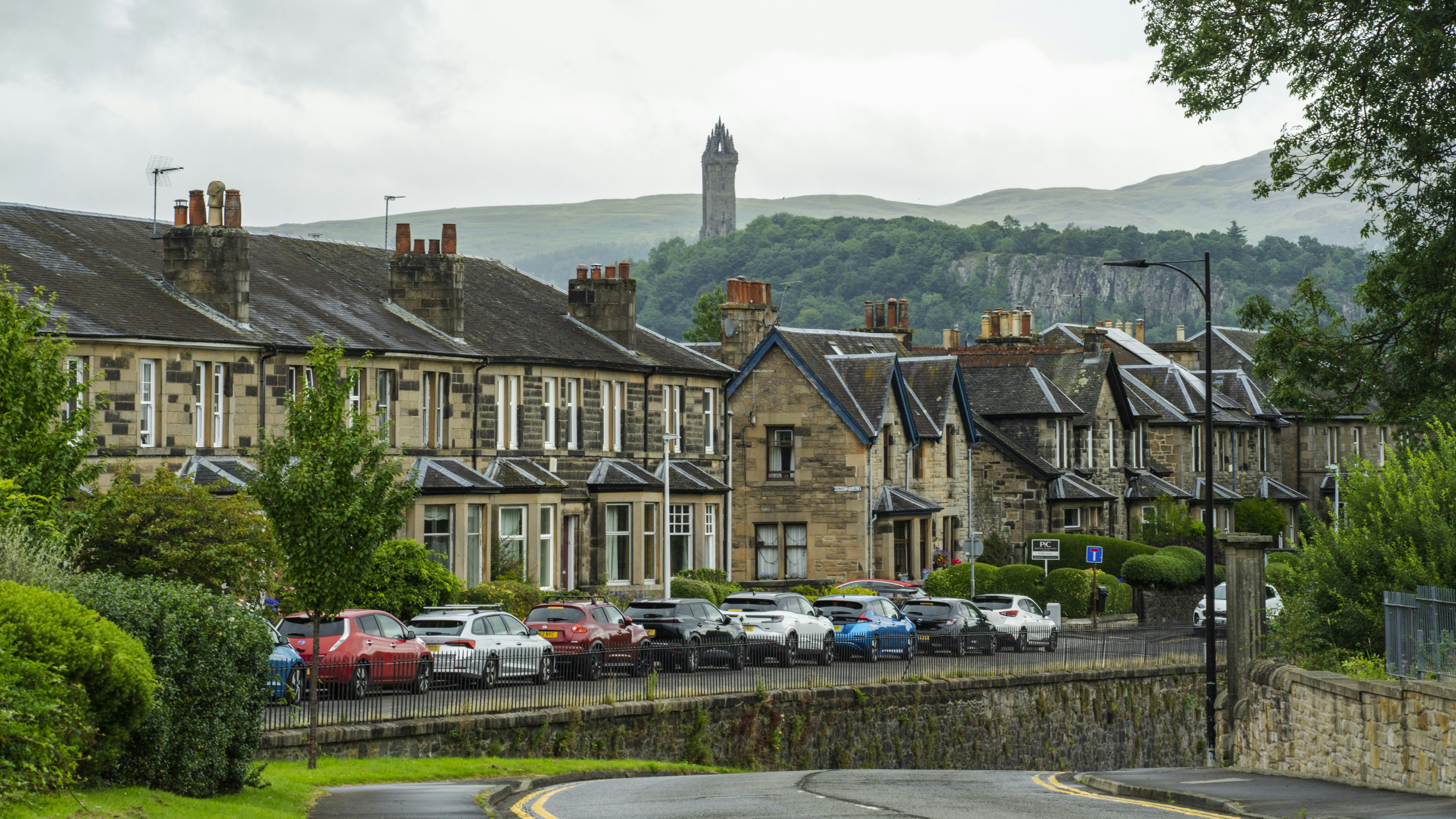 Victorian-style houses line a street with parked cars, while a historic tower looms in the background against a misty hillside.