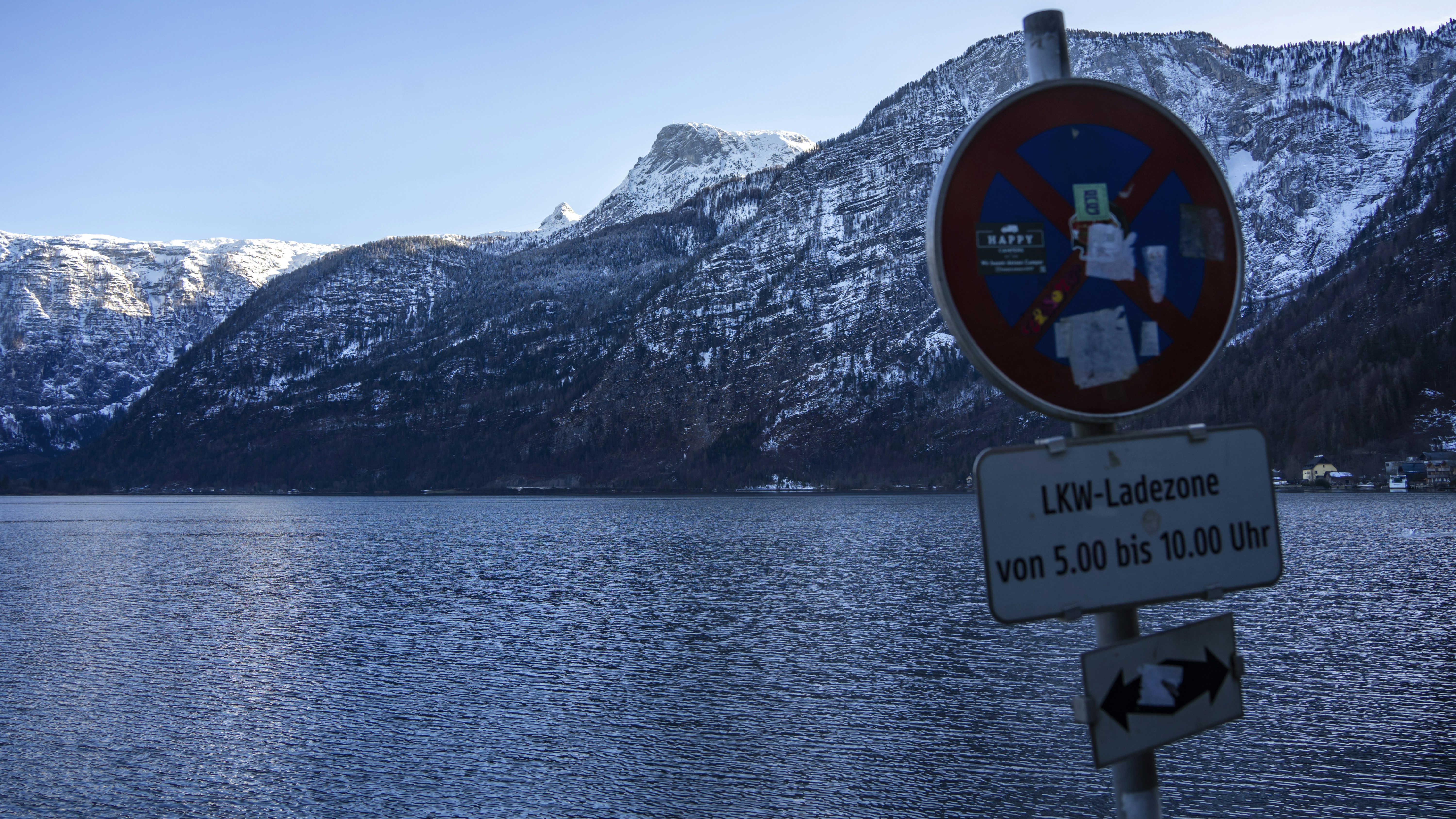 A no-parking sign stands prominently by a serene lake, framed by snow-capped mountains under a clear sky.