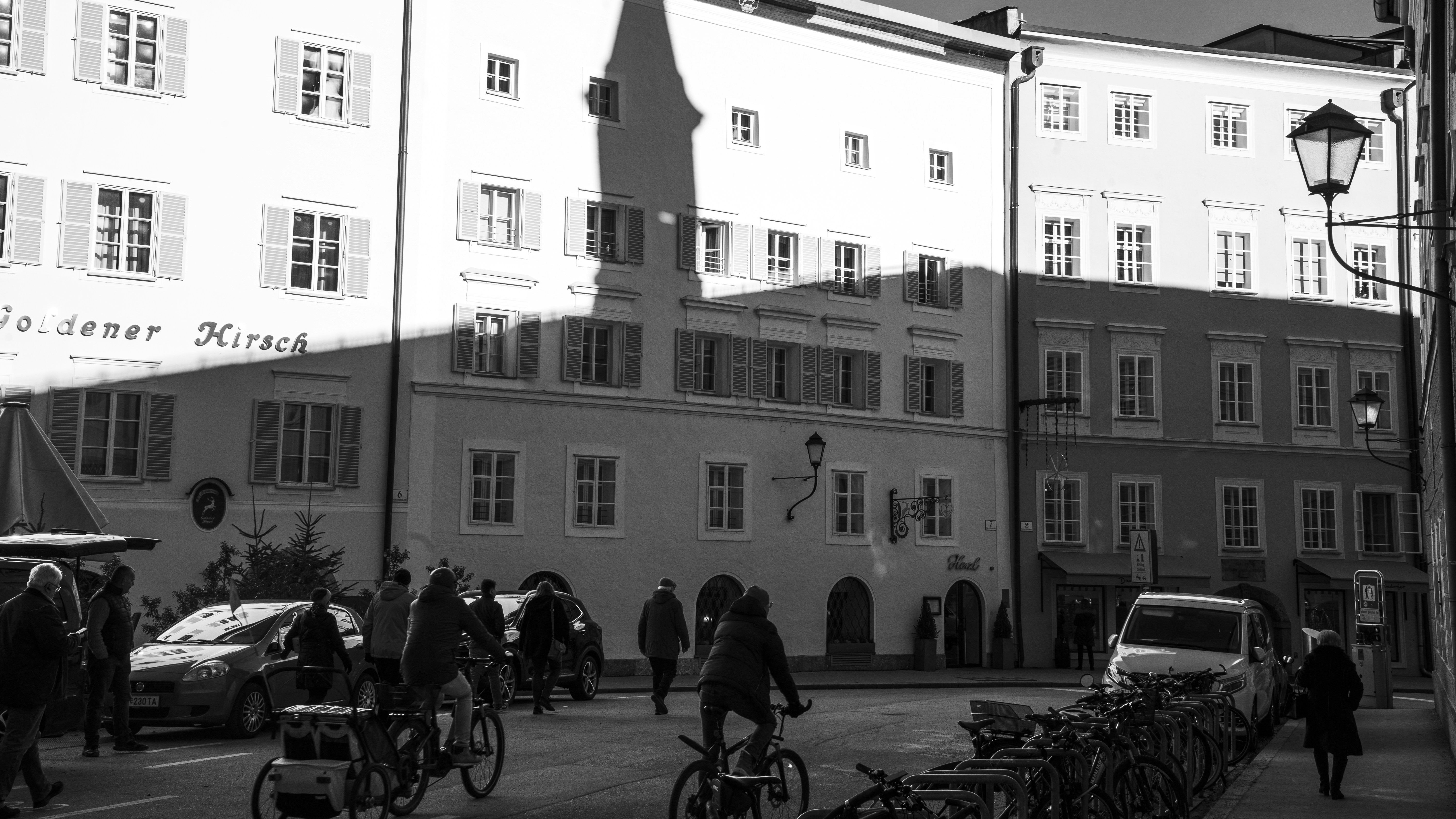 A monochrome scene depicting cyclists and pedestrians navigating a sunlit street, with shadows cast dramatically on historic buildings.
