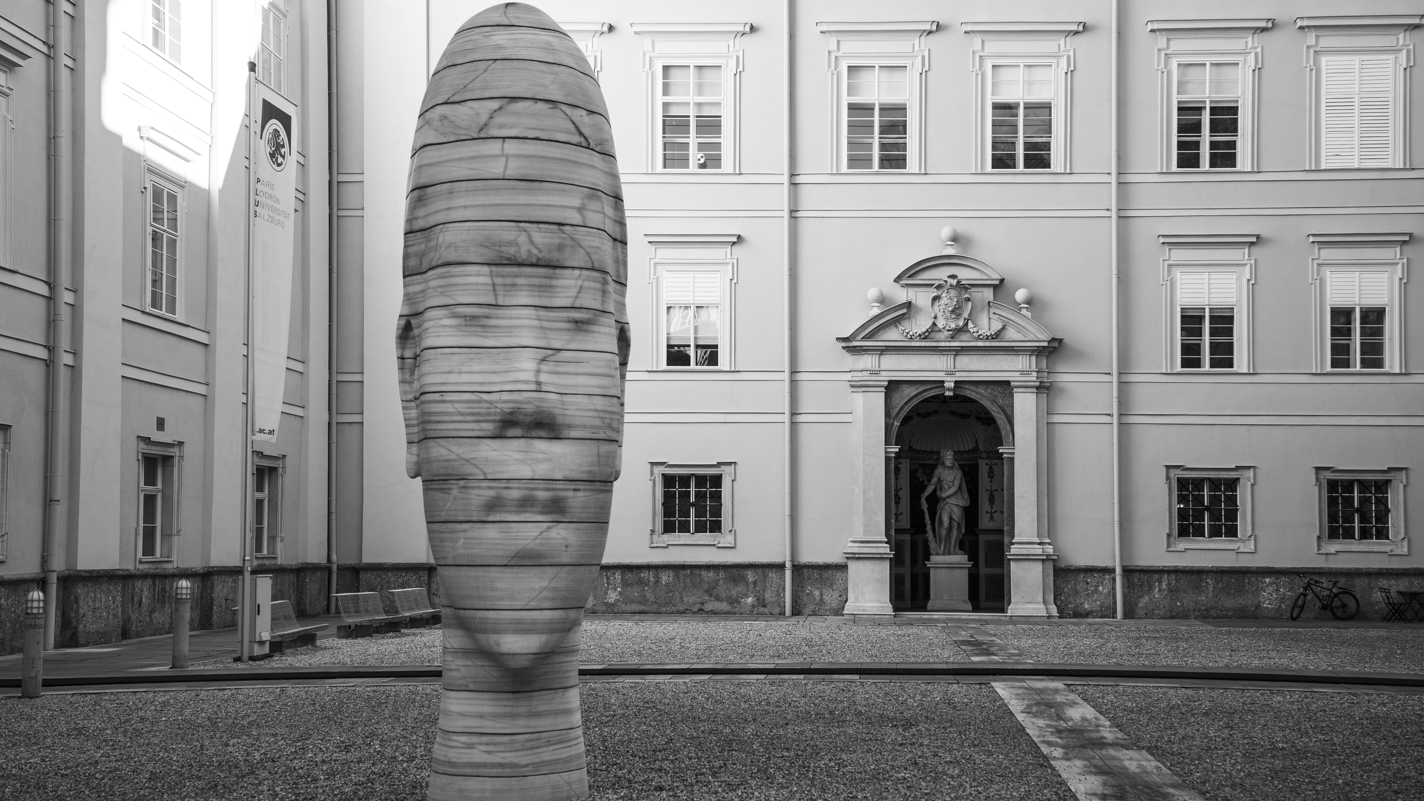 Abstract sculpture resembling a human face stands in a courtyard, framed by classic architecture. The monochrome palette enhances the interplay of light and shadow.