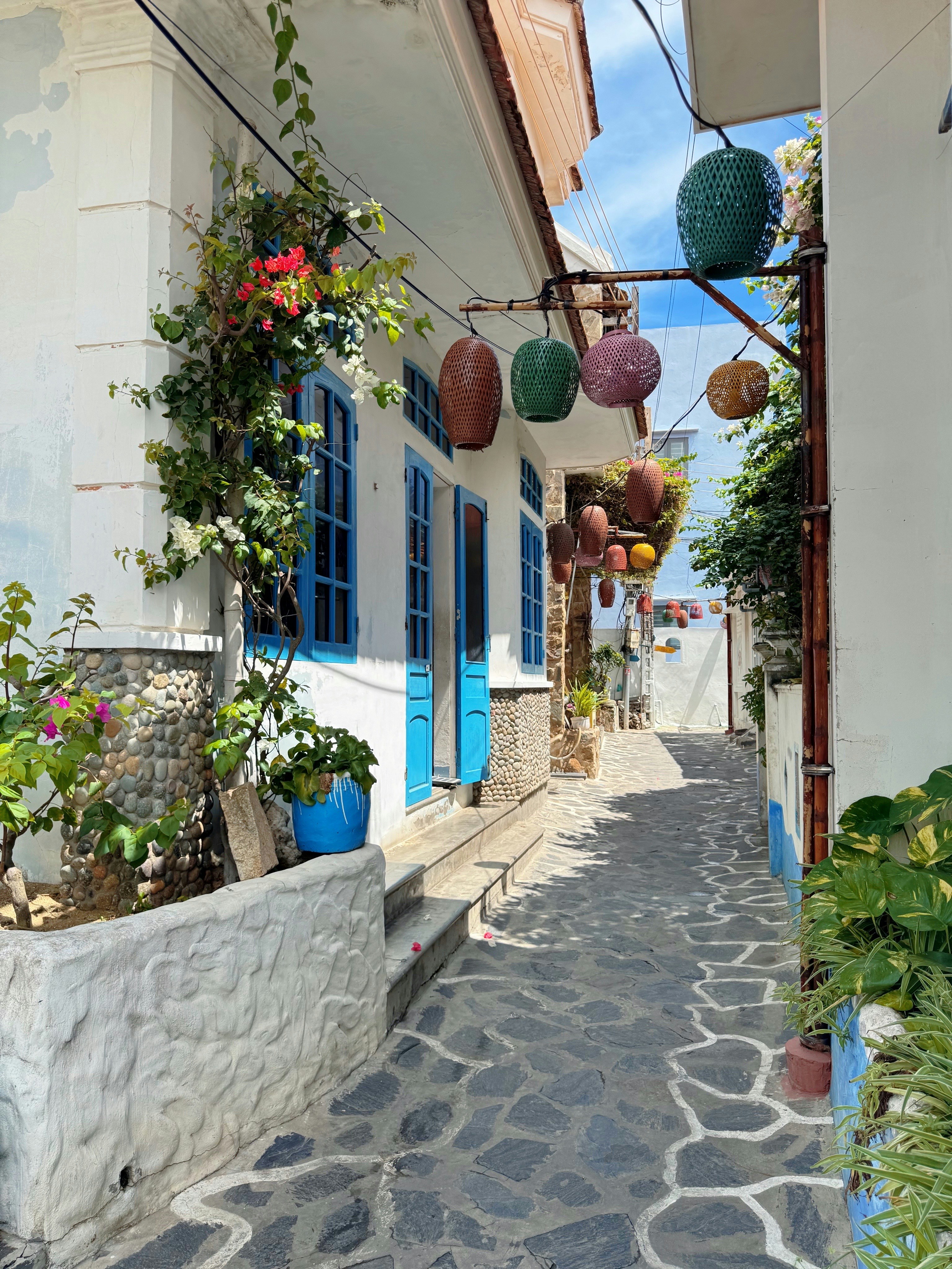 Narrow cobblestone alleyway with colorful lanterns and flowers.